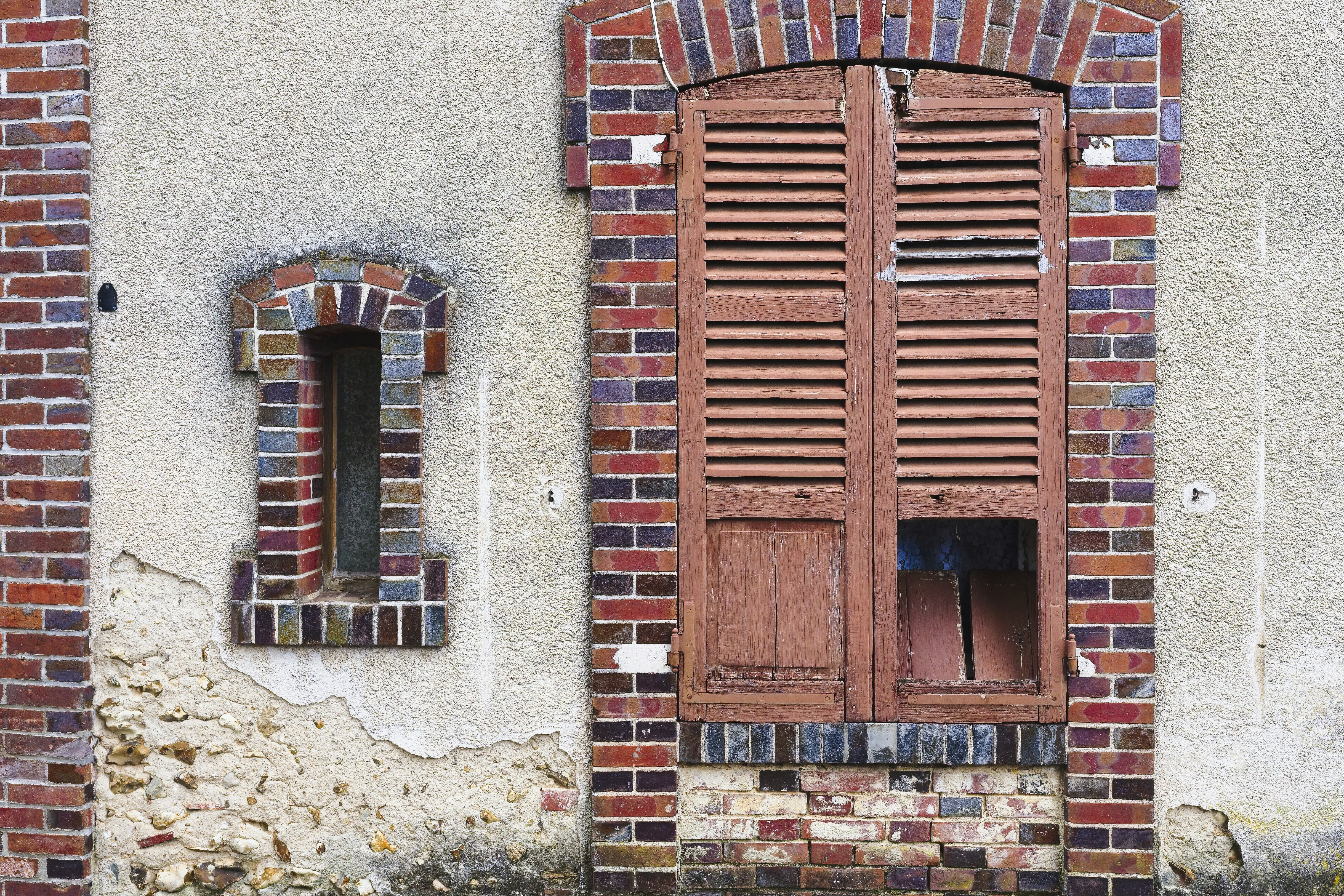 Brick Wall With Window Shutters and Small Window in France - A weathered brick wall features a large window with wooden shutters and a small arched window below. The wall is cracked and crumbling in parts.