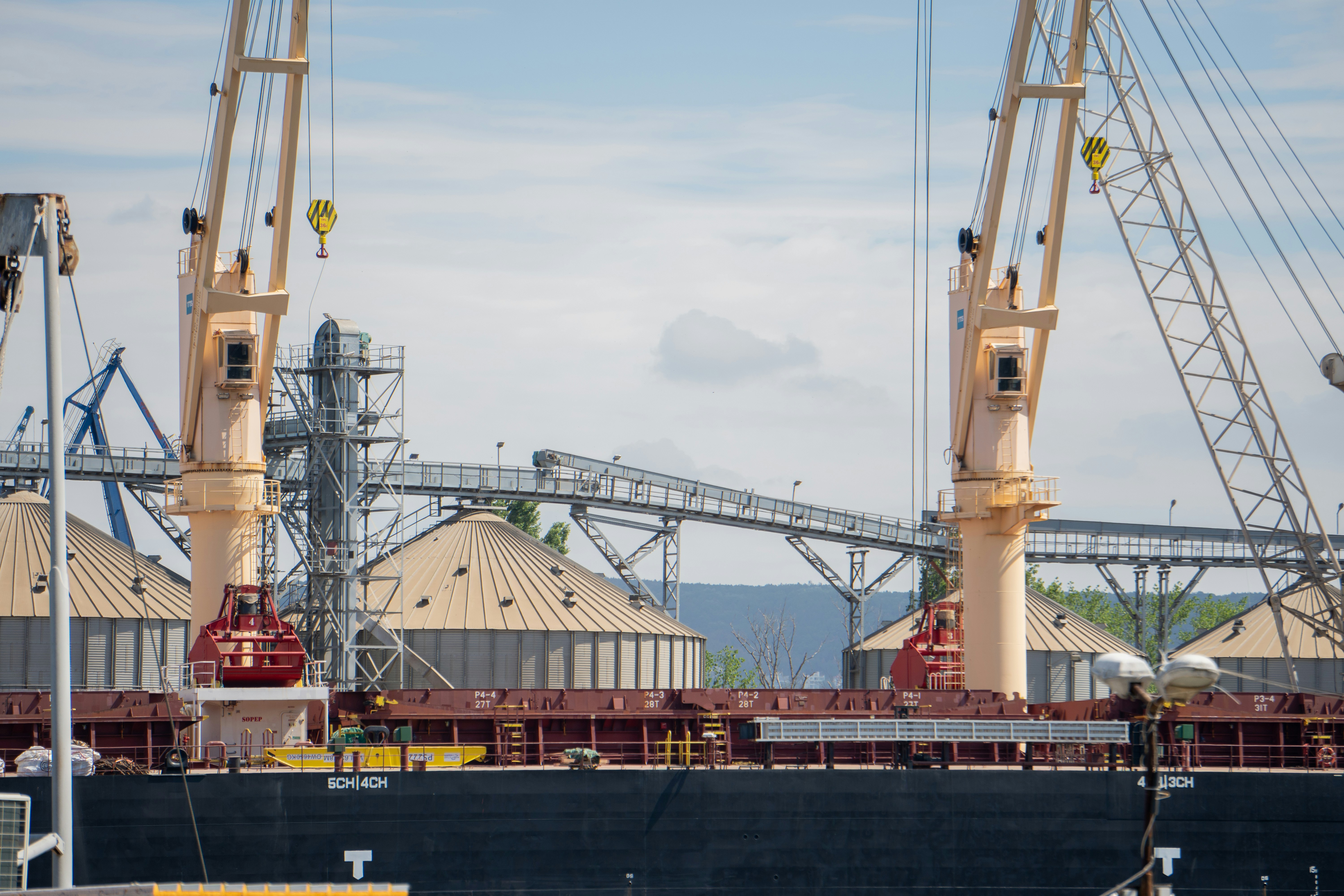 Cargo ship docked at an industrial port with large cranes and silos in the background under a cloudy sky.