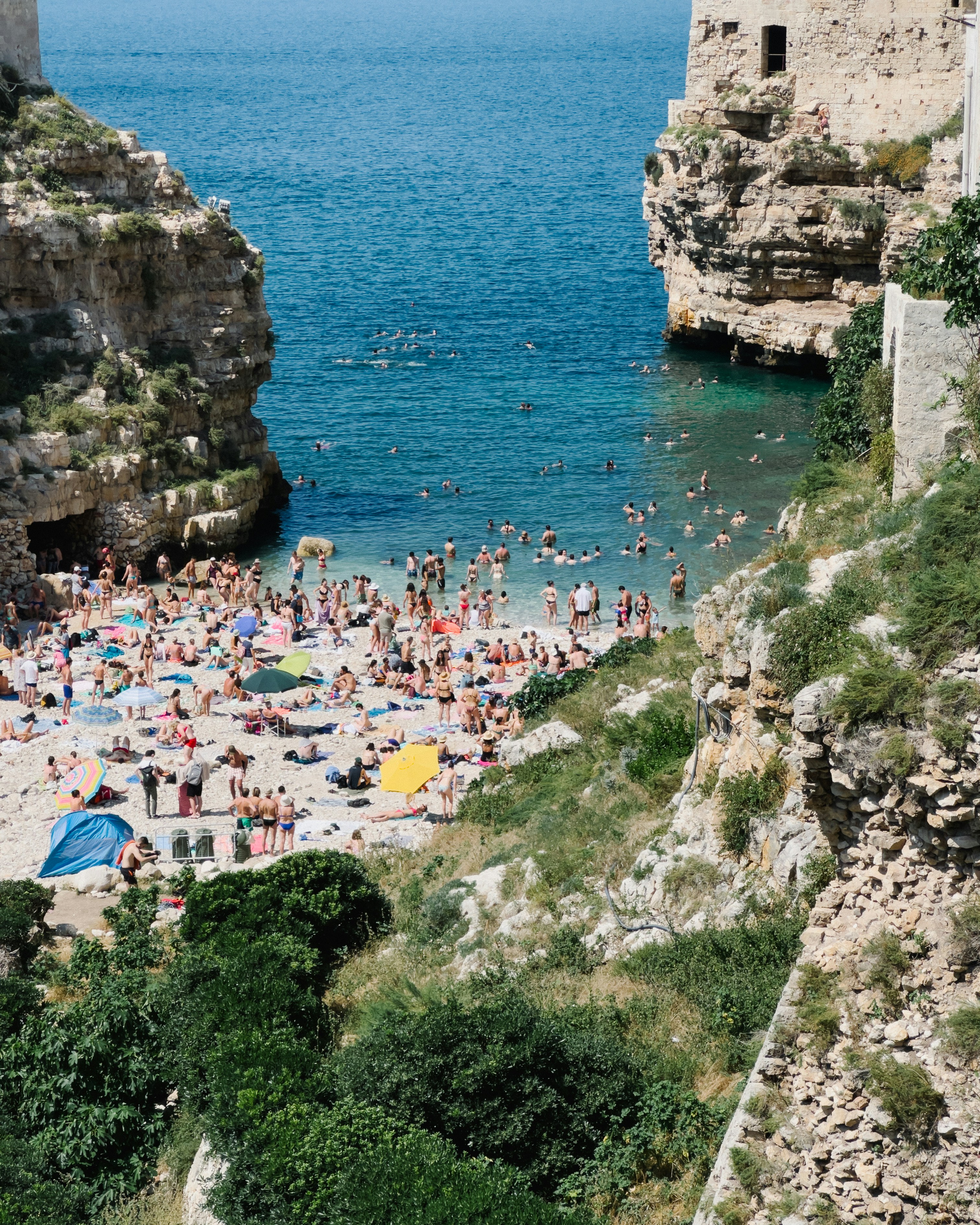 Cliffside beach tucked between limestone cliffs along a turquoise cove. Sunbathers, umbrellas, and swimmers fill the rocky shore under bright summer light.