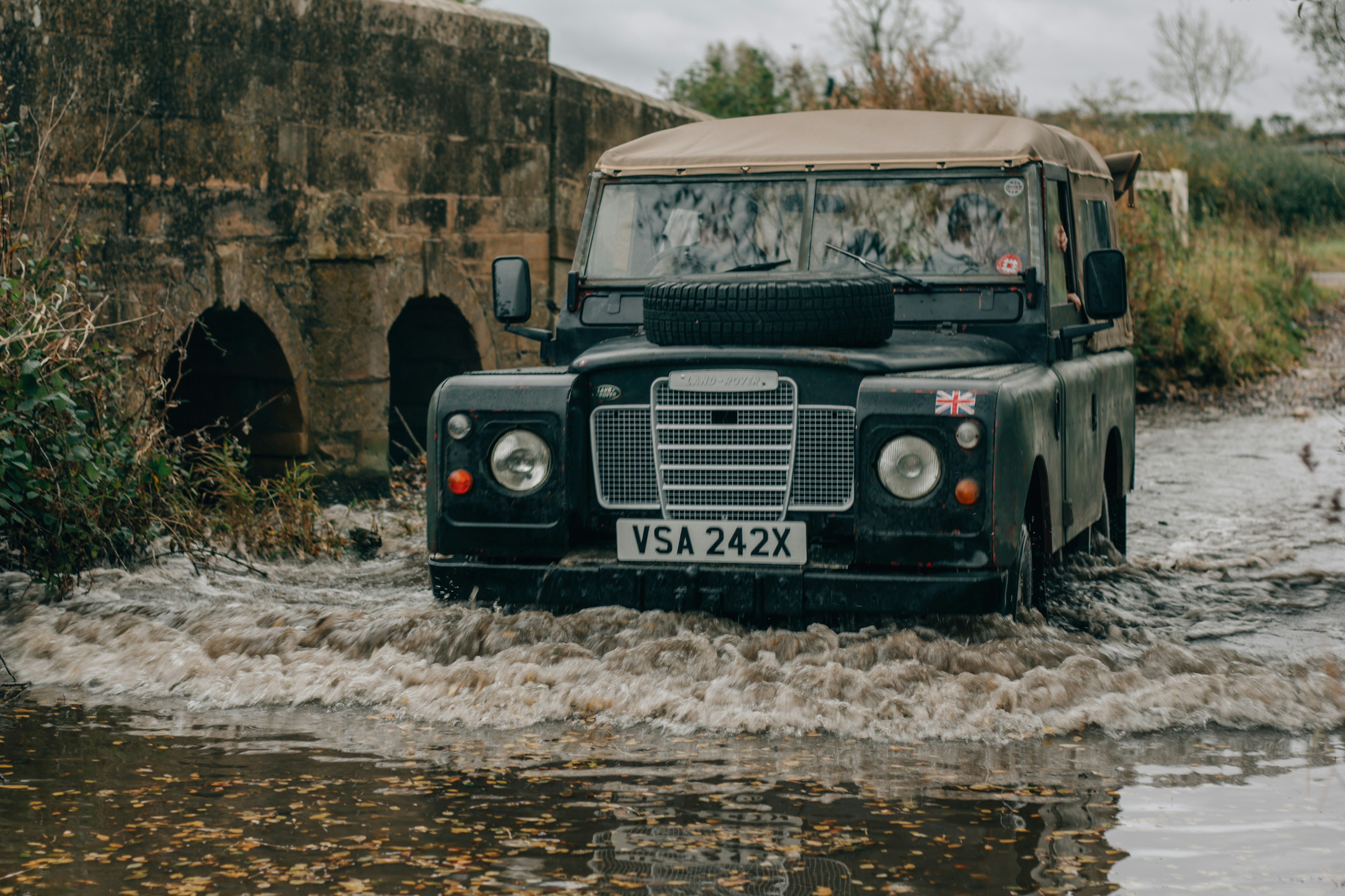 Mud-Splashed Defender Crosses a Flooded Ford Beneath a Weathered Stone Bridge