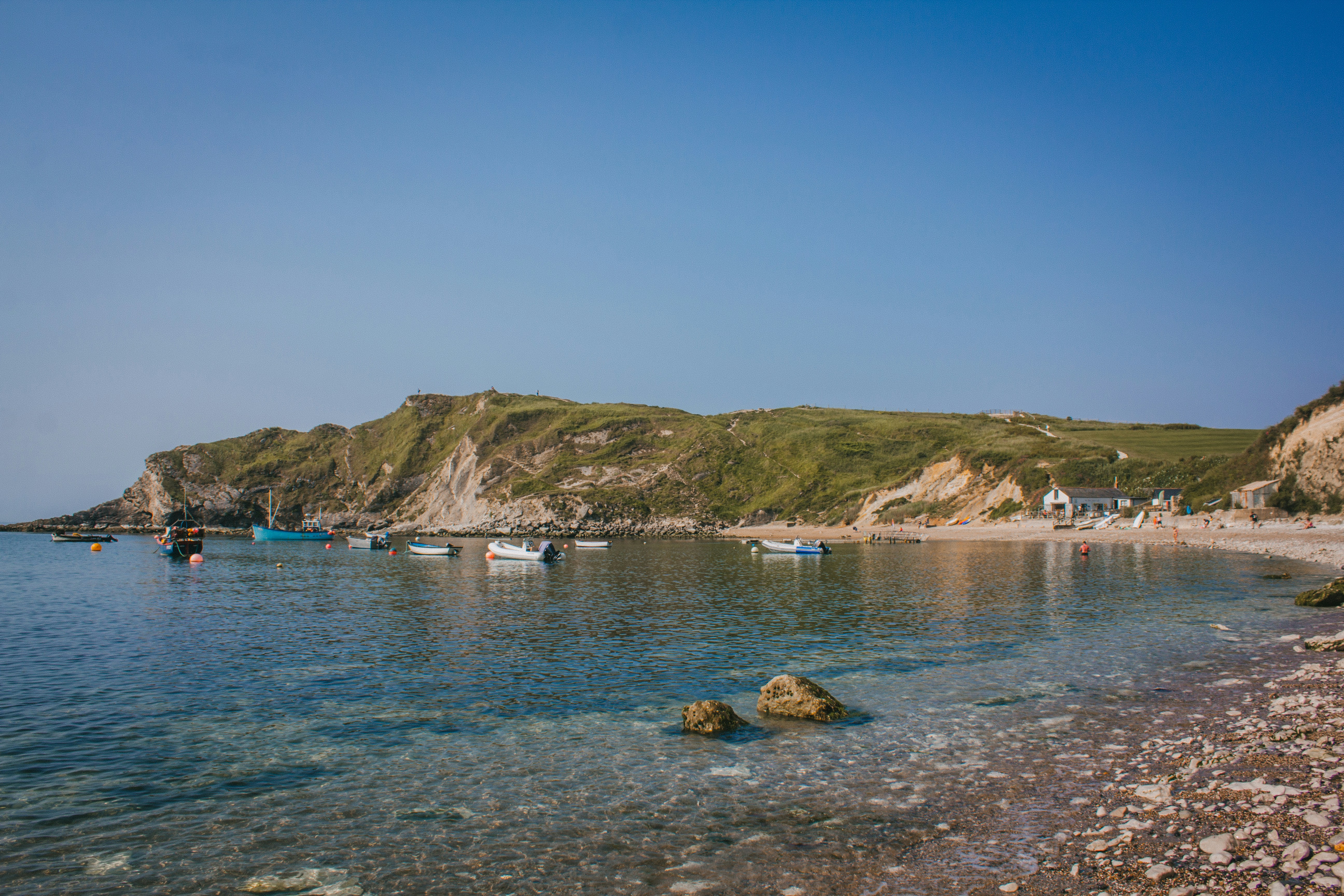 Tranquil beach scene featuring boats anchored near a rocky coastline under a clear blue sky.
