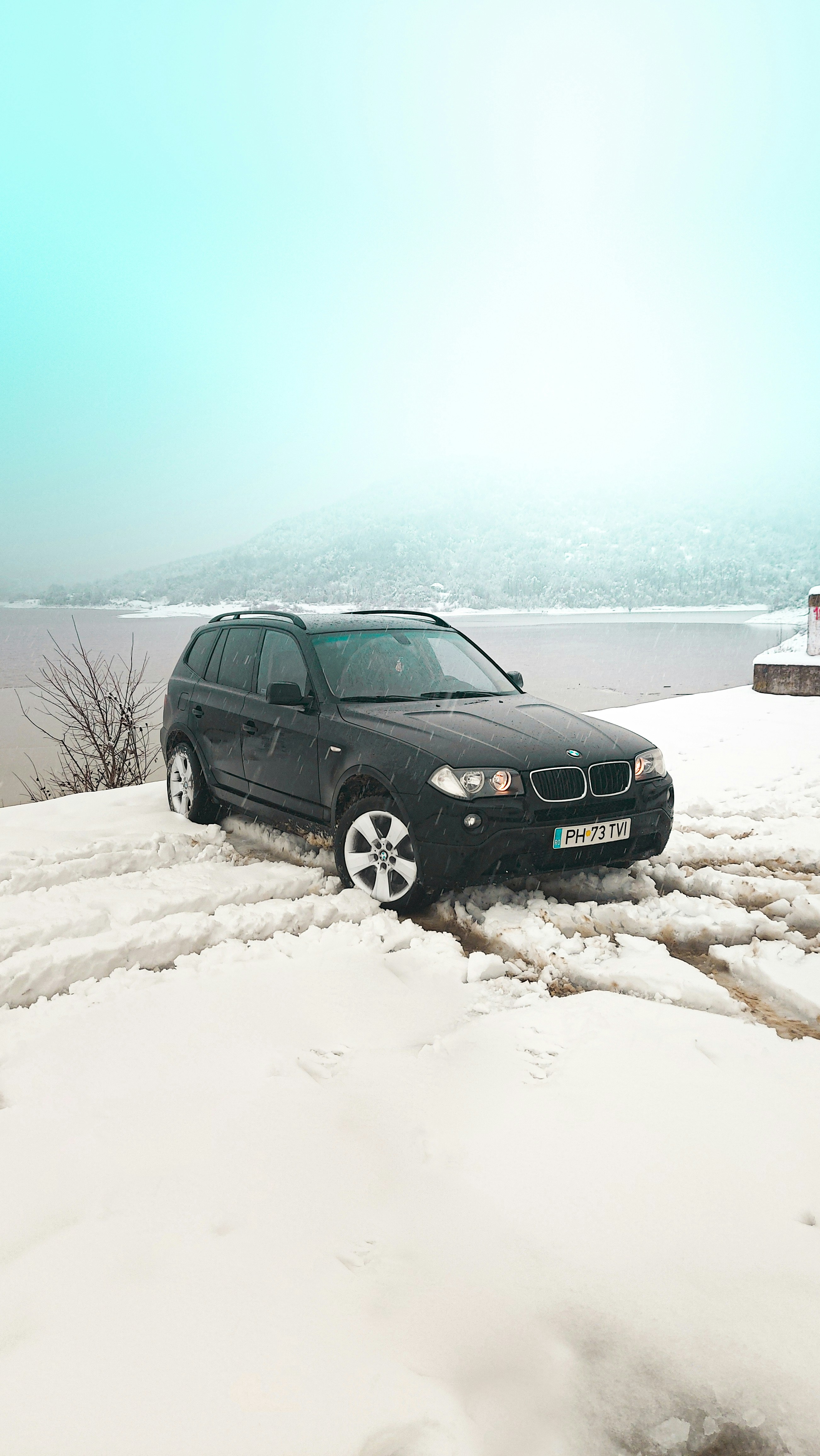 Dark BMW SUV parked in deep snow along a lakeshore, with misty hills and a pale blue sky.