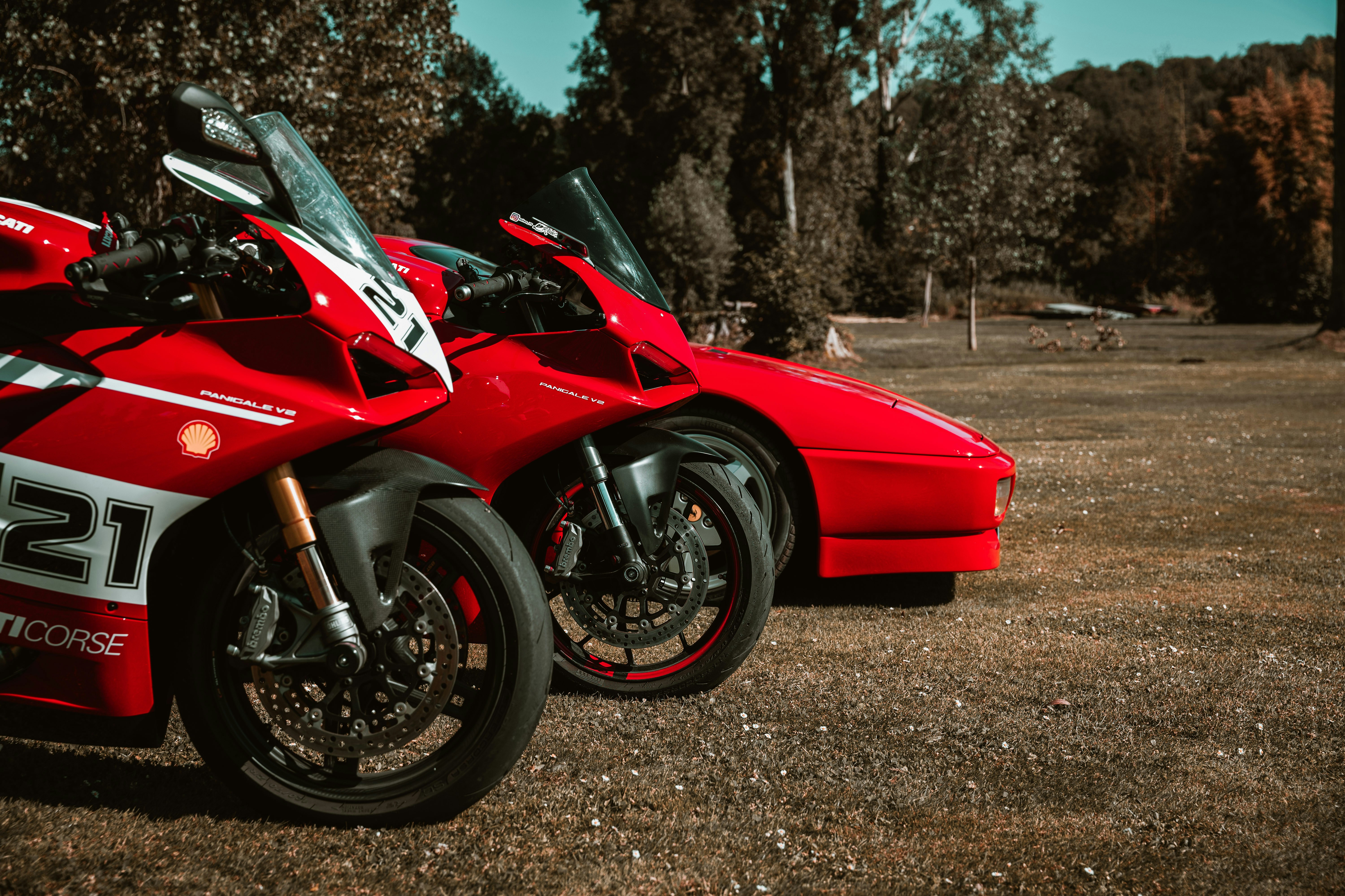Two iconic red vehicles, a Ducati motorcycle and a Ferrari sports car, showcased side by side on a sunny day in a lush green setting.