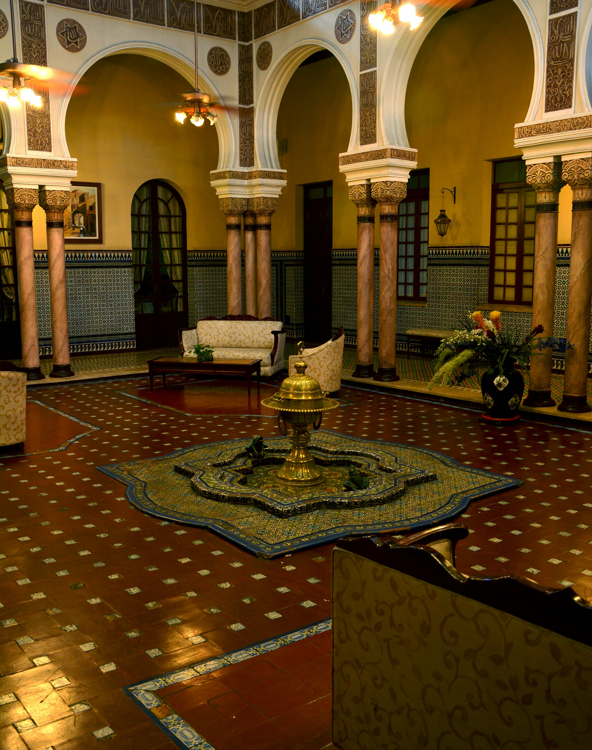 Intricate Moroccan-style interior featuring ornate pillars, a decorative fountain, and plush seating arrangements. The warm ambiance invites relaxation and reflection.