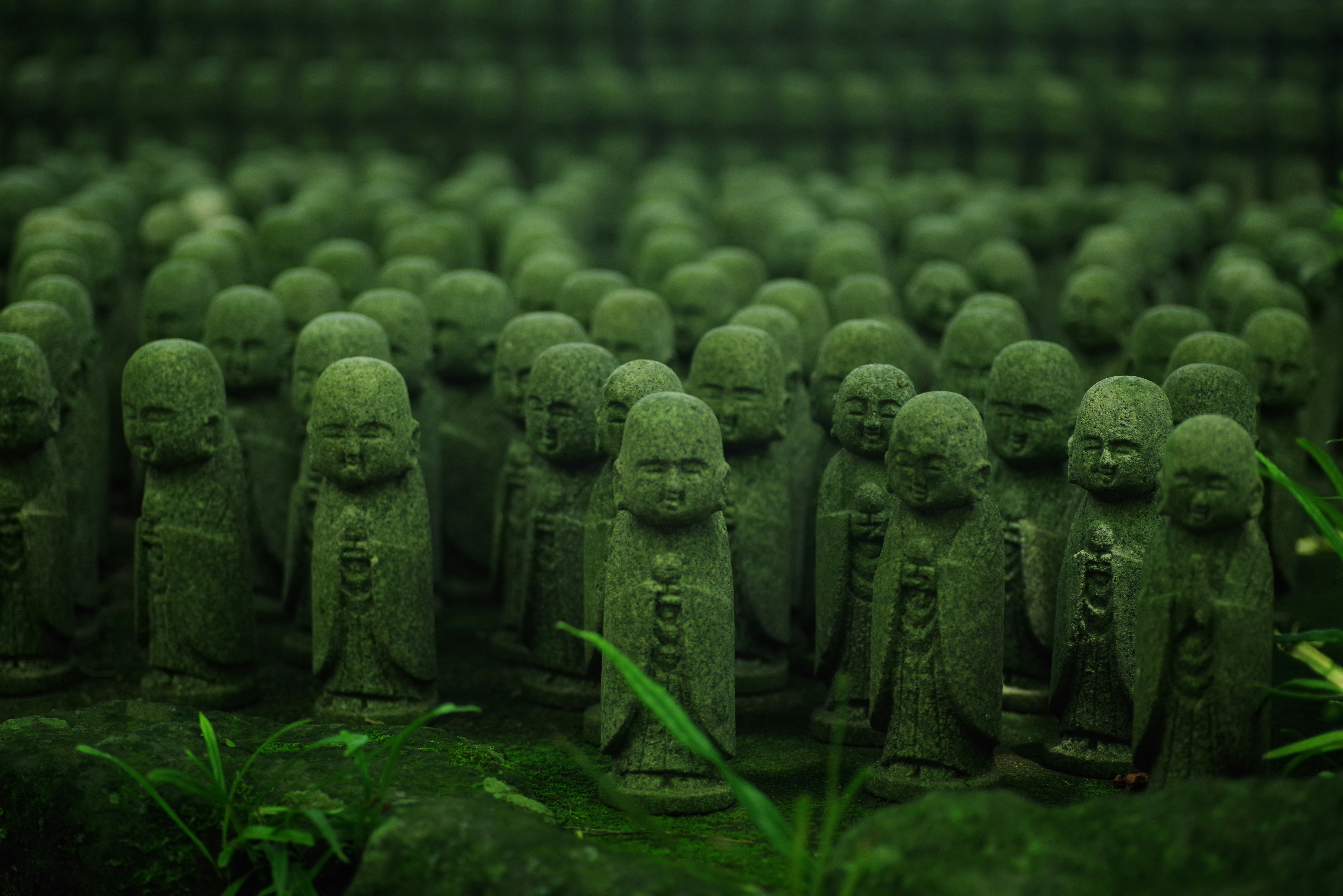 Moss-covered stone monks stand in orderly rows within a dim, green-lit garden. The scene emphasizes quiet, meditative stillness.