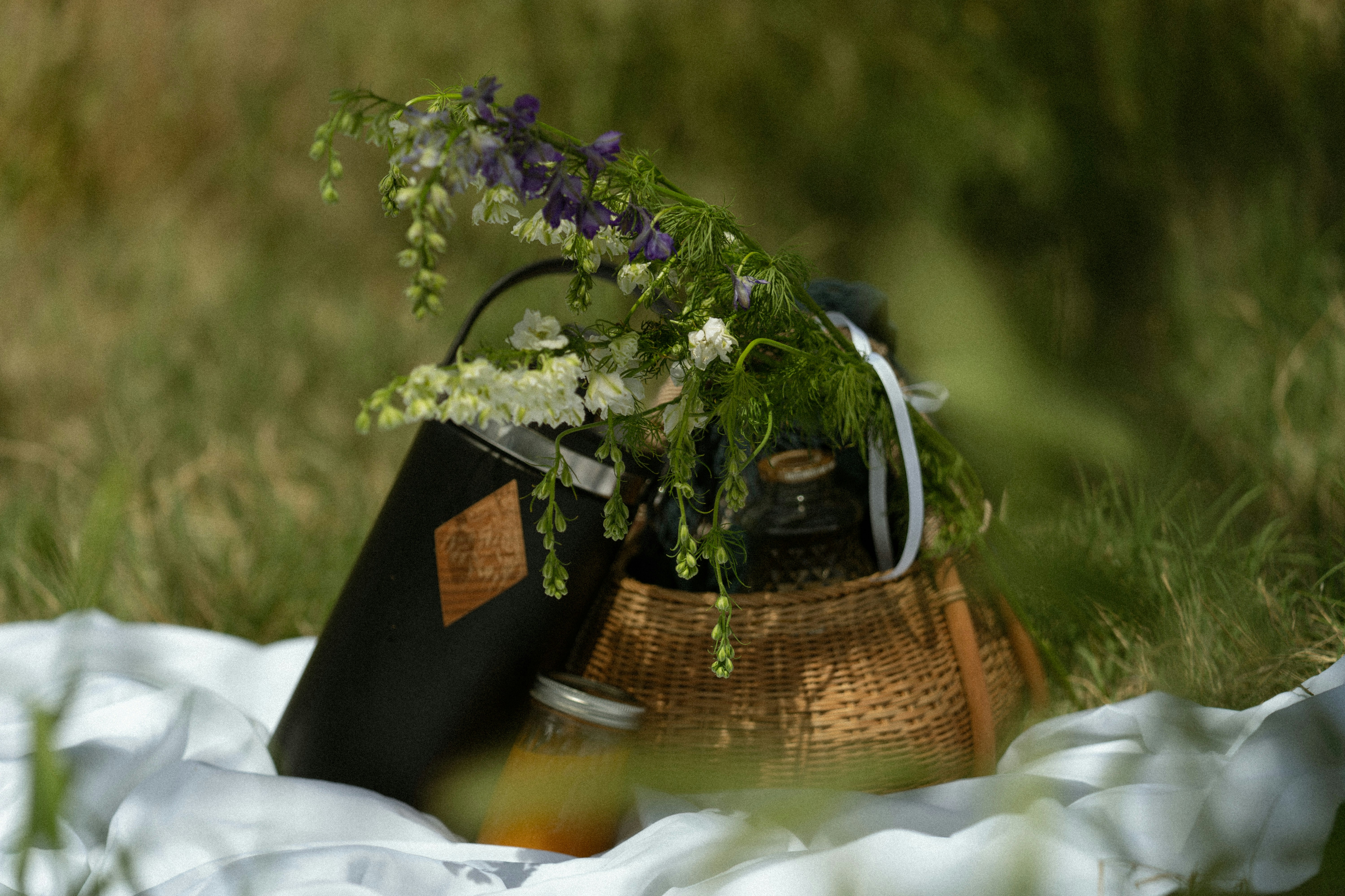A woven basket filled with fresh flowers and a vintage container rests on a white cloth in a grassy setting.