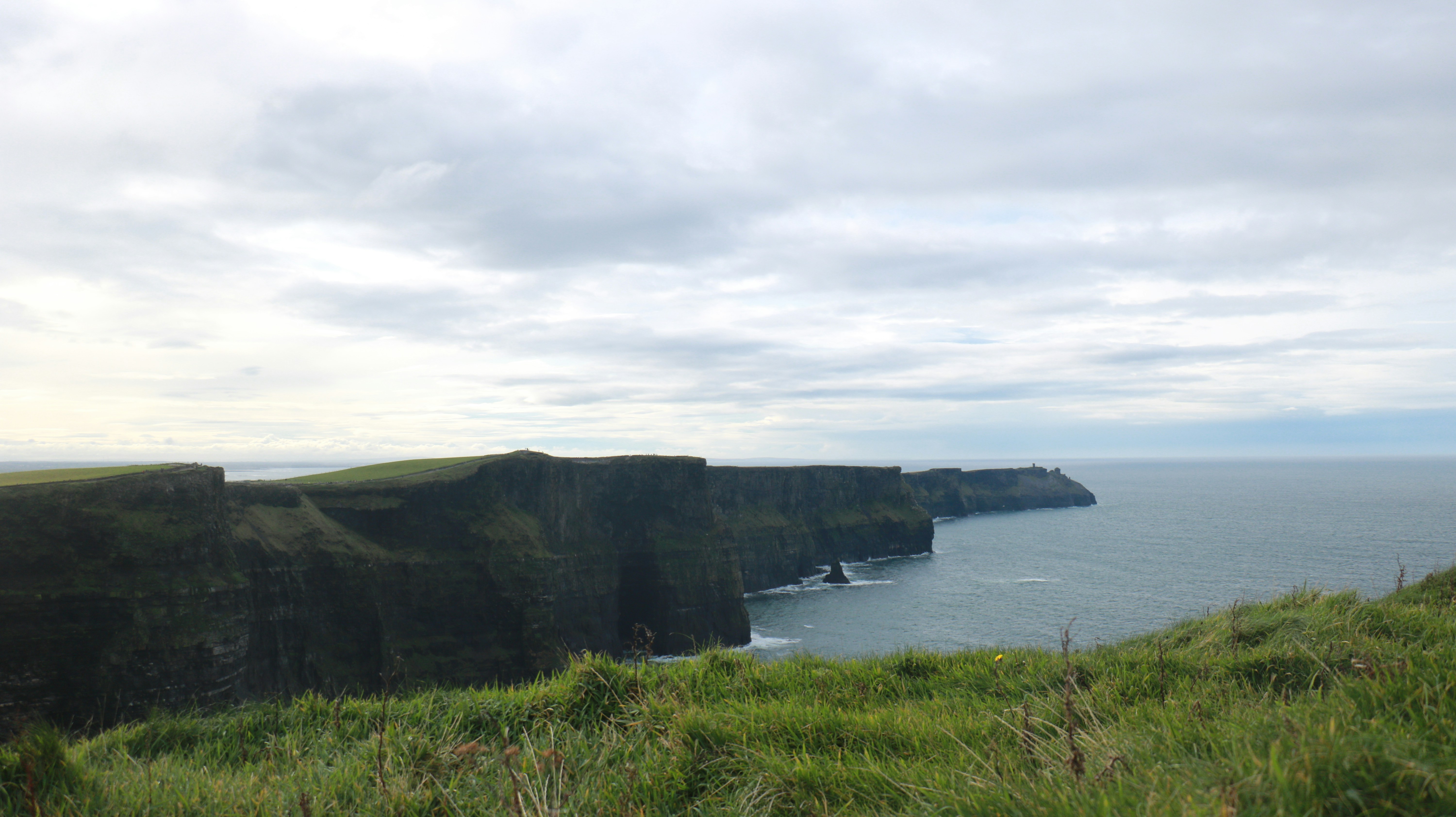 Majestic cliffs rise dramatically from the Atlantic Ocean, framed by lush greenery under a cloudy sky. The scene captures the rugged beauty of the coastline.