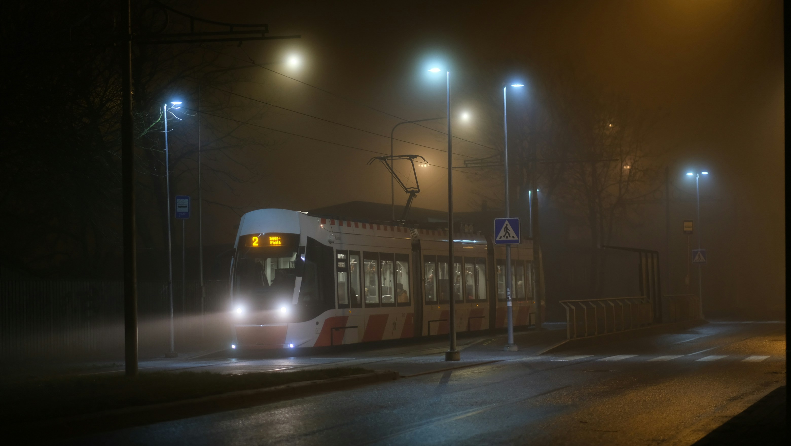 A tram in Kalamaja at night. The tram is illuminated by streetlights, creating a misty, atmospheric scene. The area is dimly lit, with the tram’s headlights cutting through the fog.