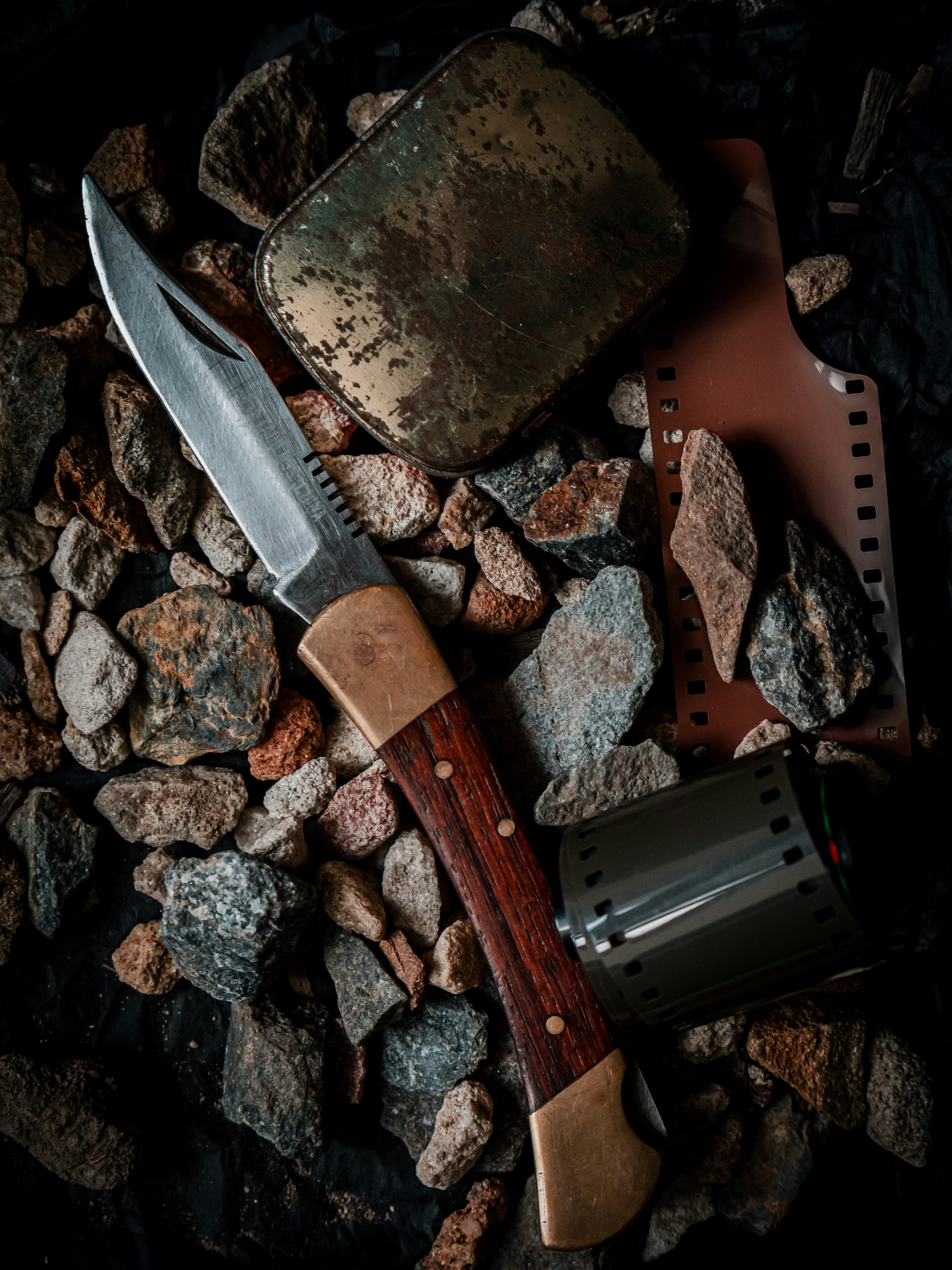 Close-up photograph of a weathered fixed-blade knife with a wooden handle nestled among rough rocks, alongside a dented metal tin and a strip of film. The composition emphasizes rugged outdoor gear and forgotten camp relics.