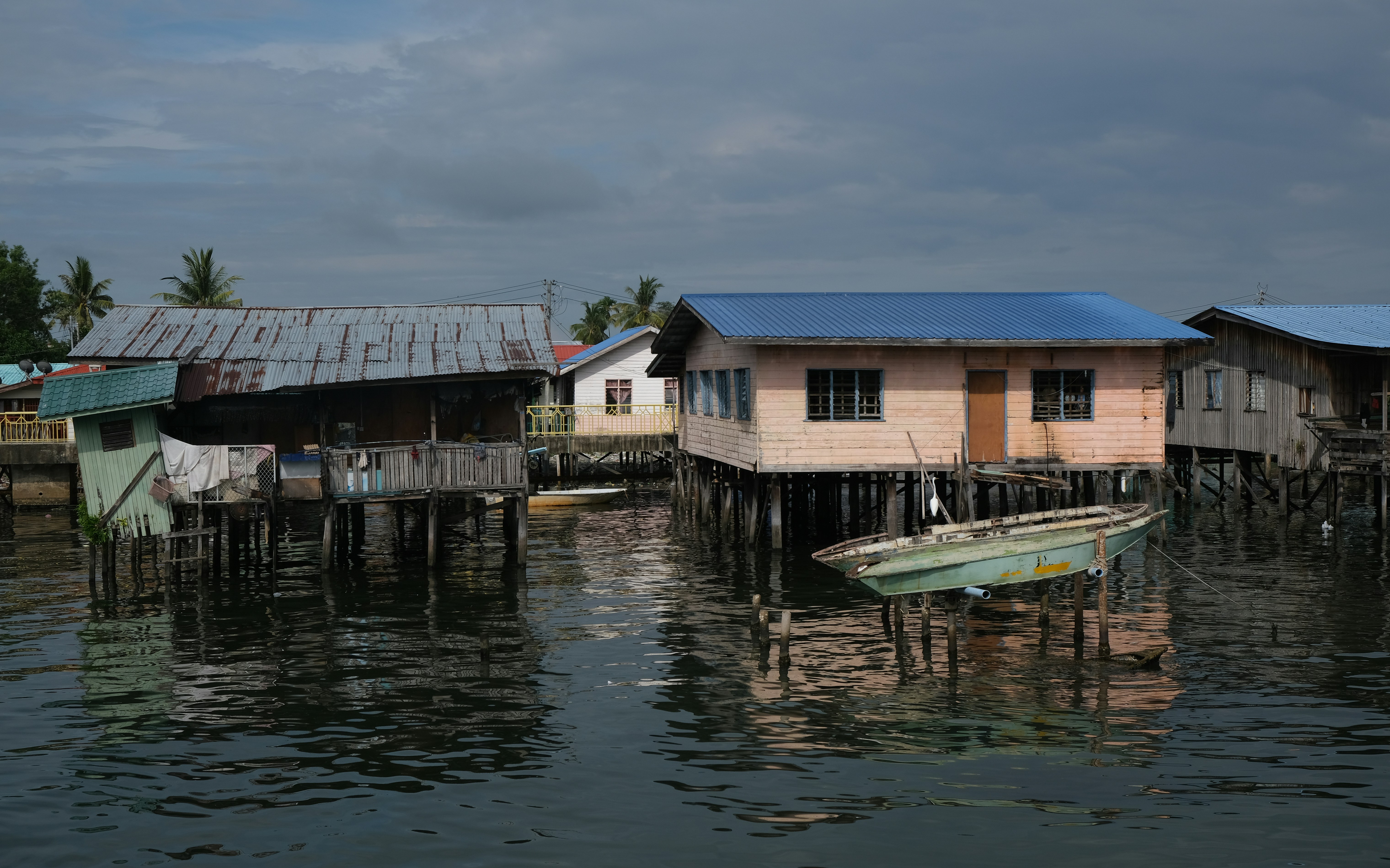 Wooden stilt houses with tin roofs rise above calm water, connected by narrow walkways under a cloudy sky.