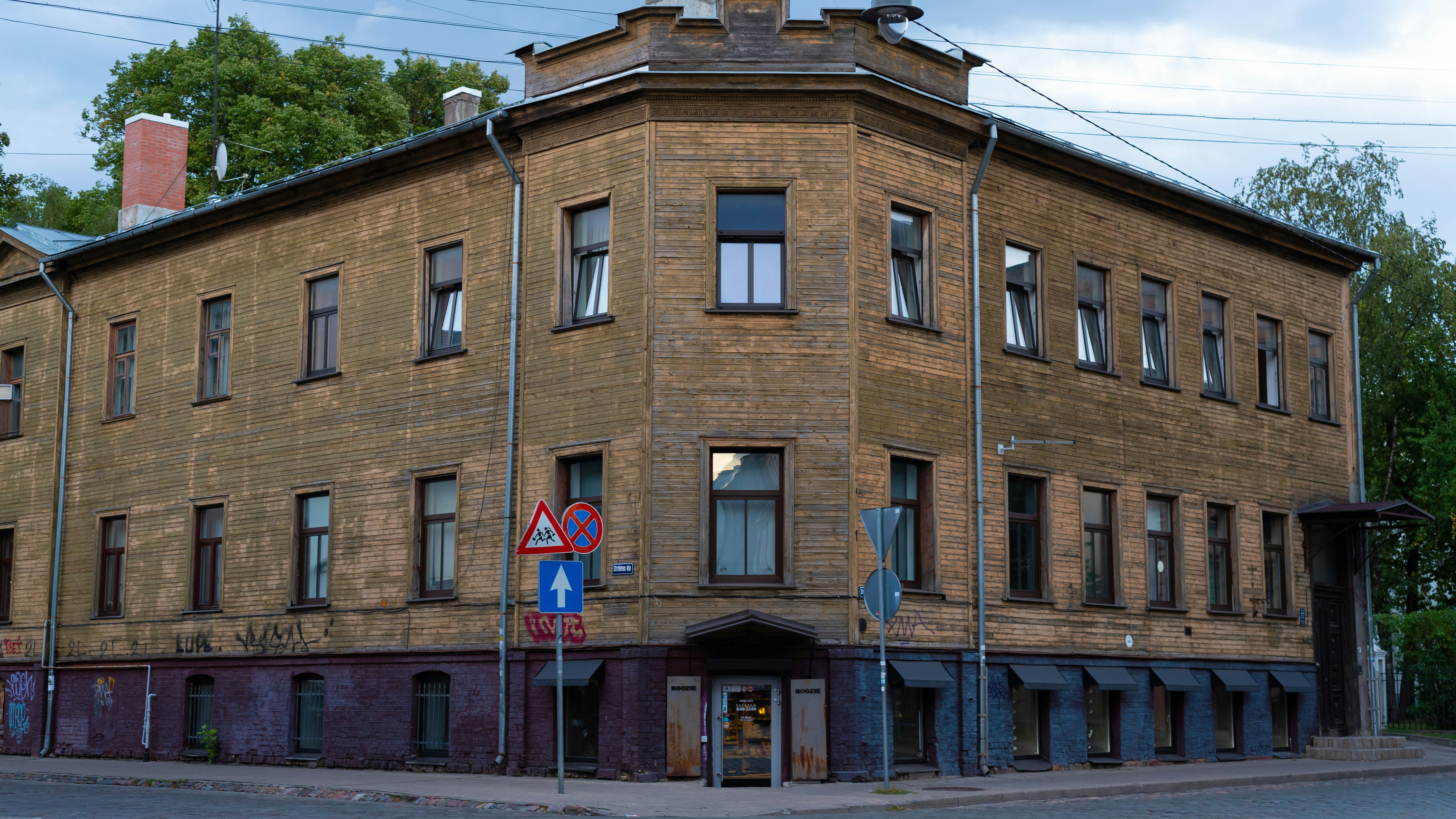 Two-story wooden building with dark brick base and evenly spaced windows, set on a quiet corner with pedestrian signage.