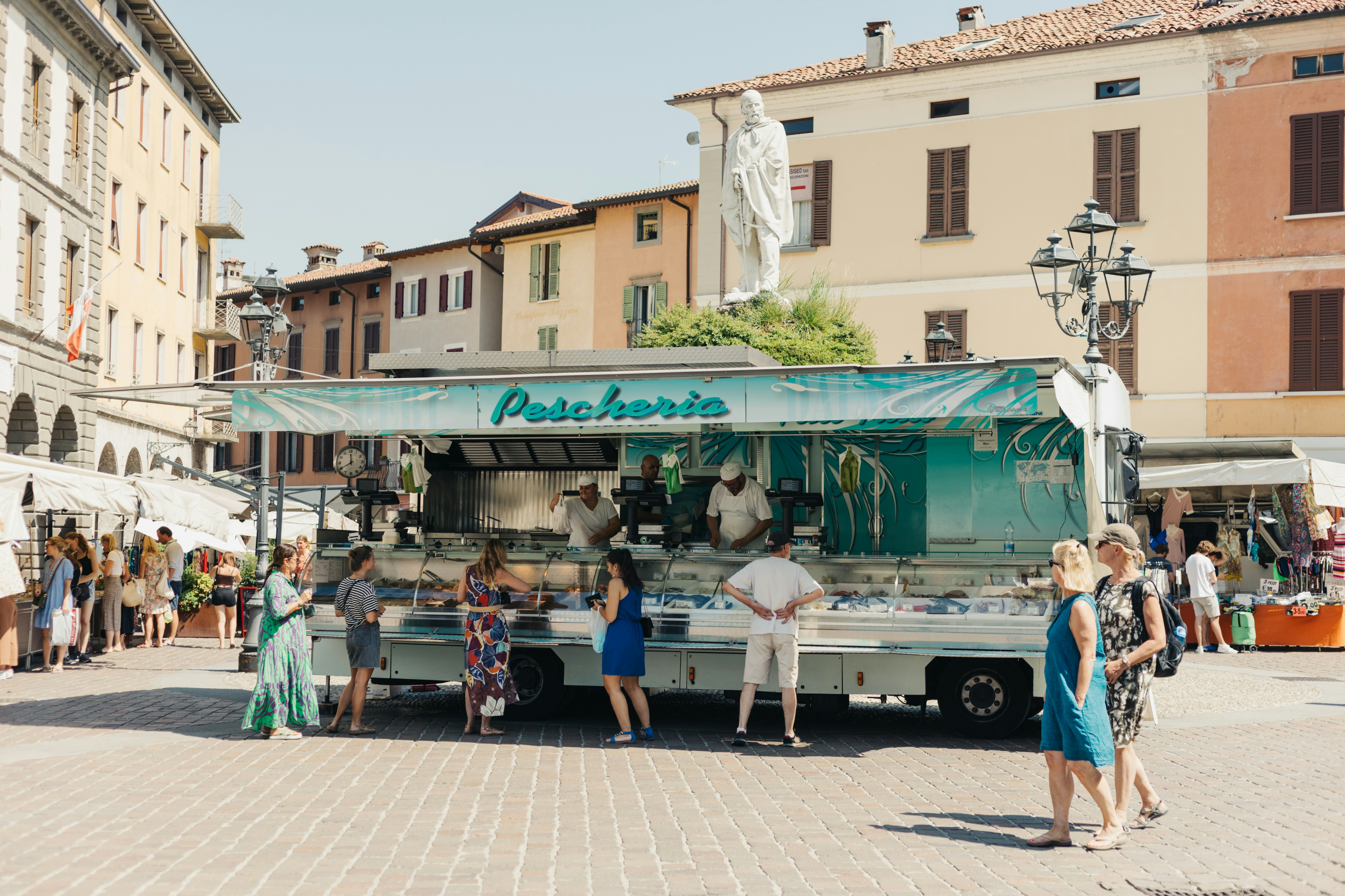 Italy in the summer, looking at a fish stand in a food market with people walking around on a sunny day.