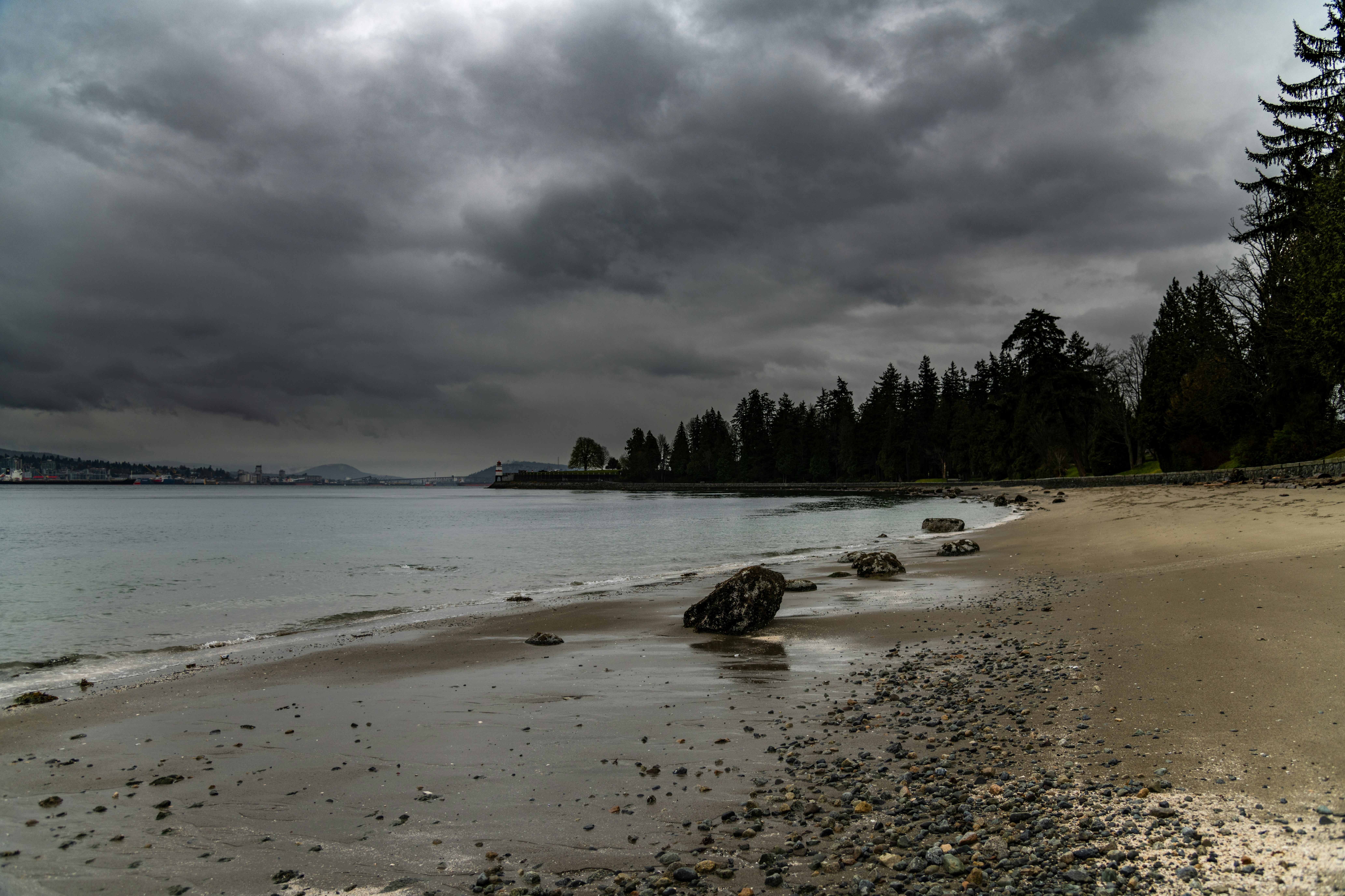 Sandy beach with pebbles facing a lighthouse, mountains, trees on a rainy day.
