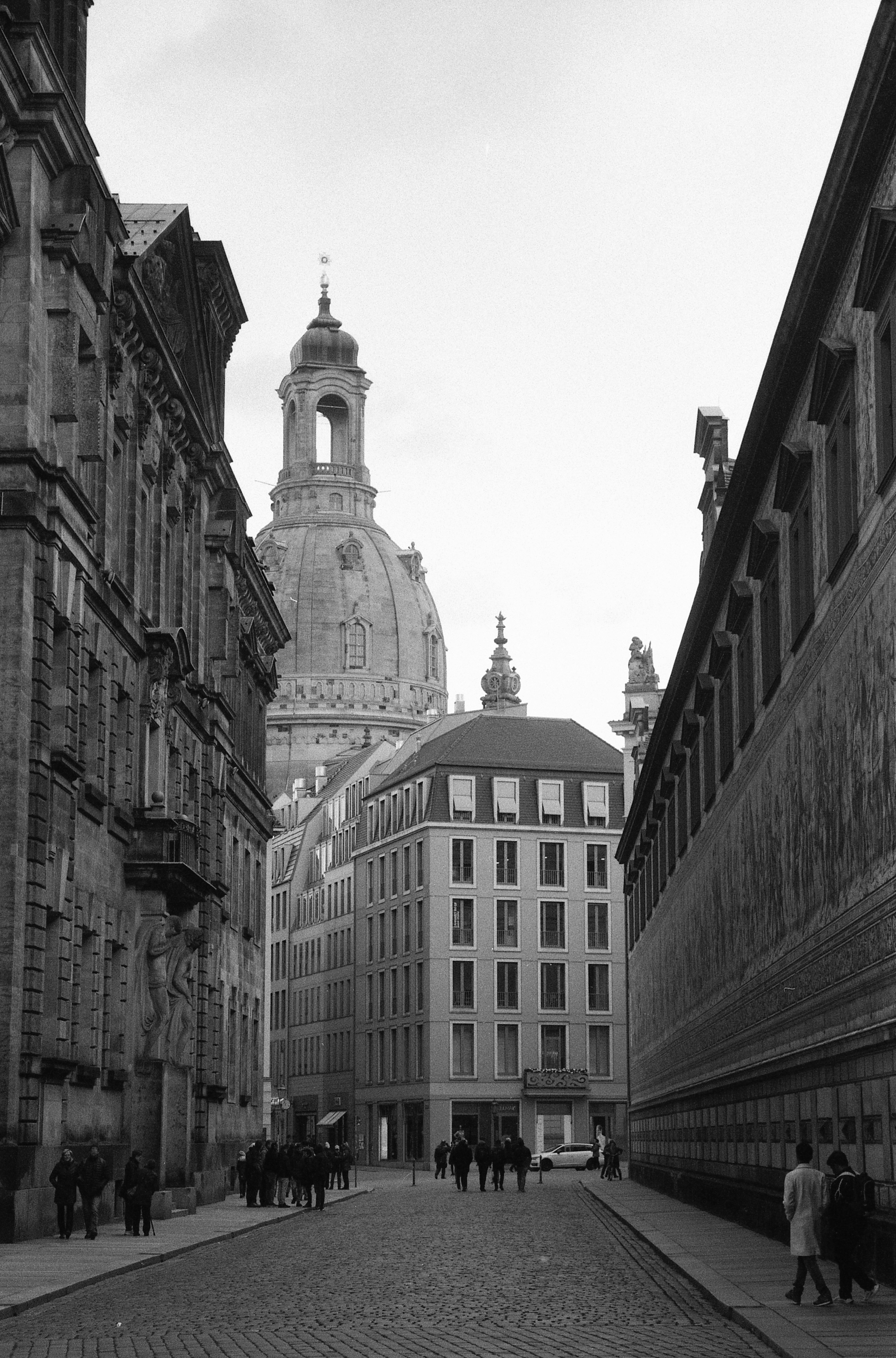 Black-and-white street scene with historic stone buildings lining a cobblestone avenue, a domed church rising behind, and pedestrians in the foreground.