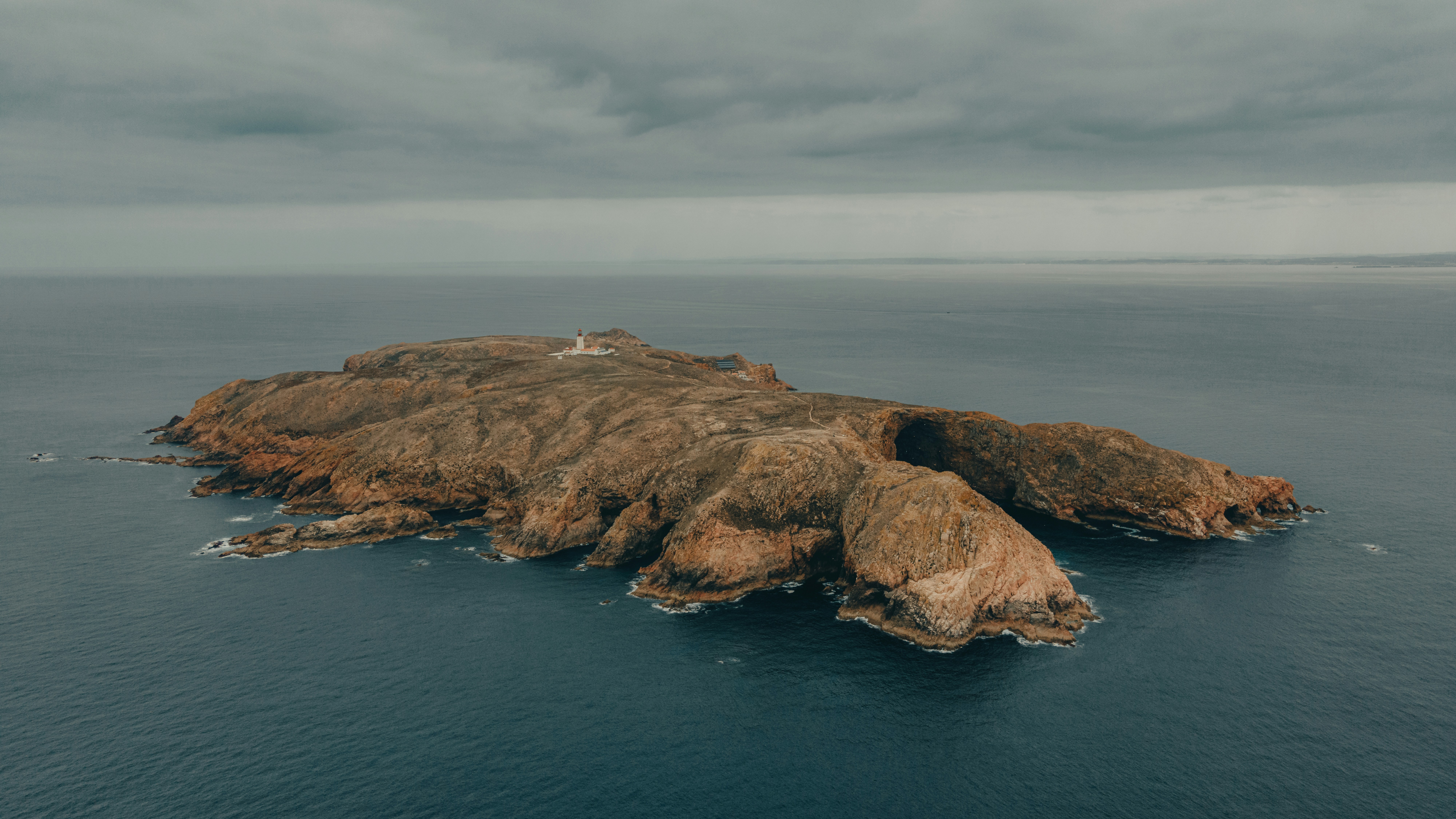 Aerial view of a rugged island surrounded by deep blue waters, featuring a distant lighthouse atop the rocky terrain.