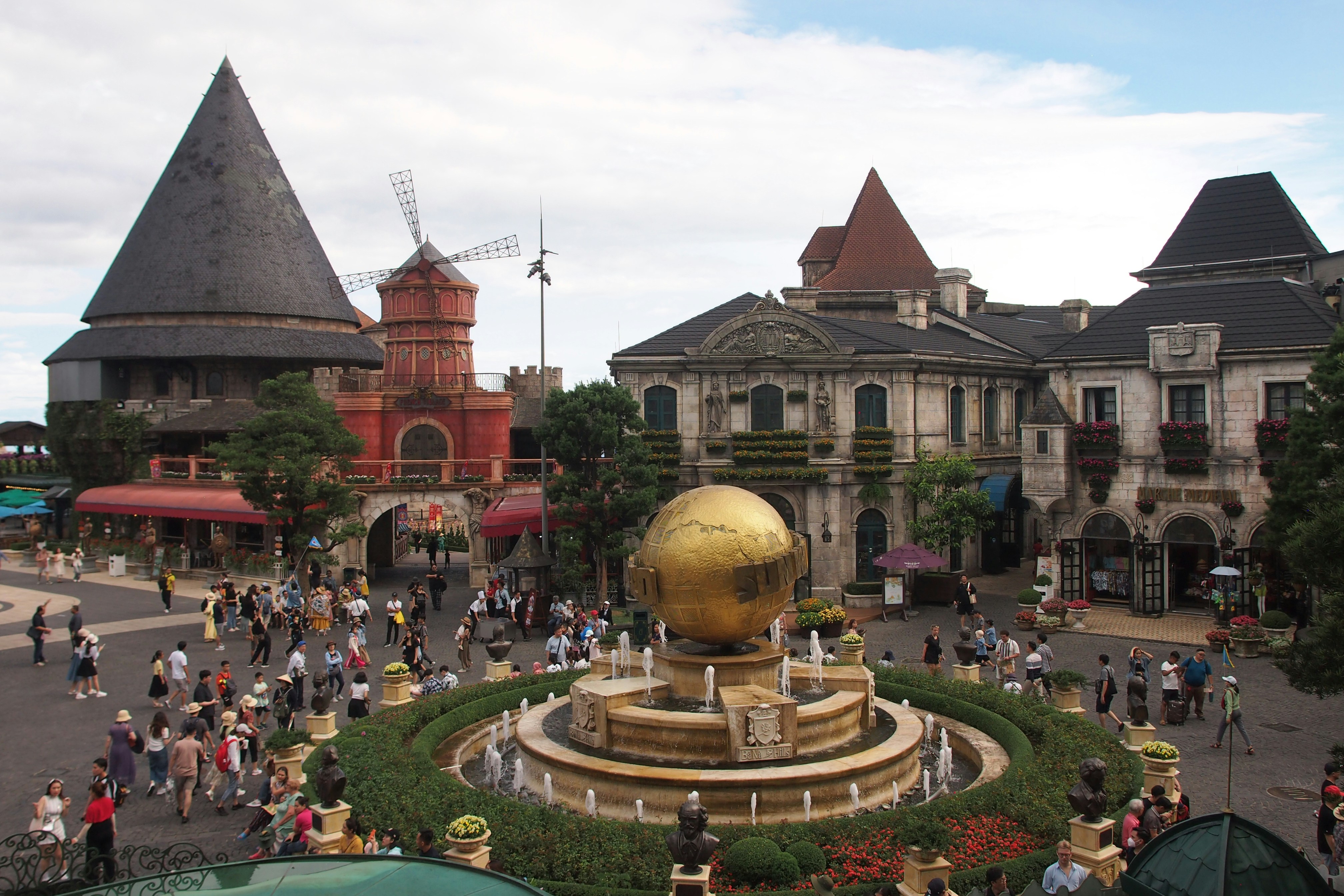 A vibrant scene in a theme park featuring a central fountain with a golden globe, surrounded by whimsical architecture and bustling visitors.