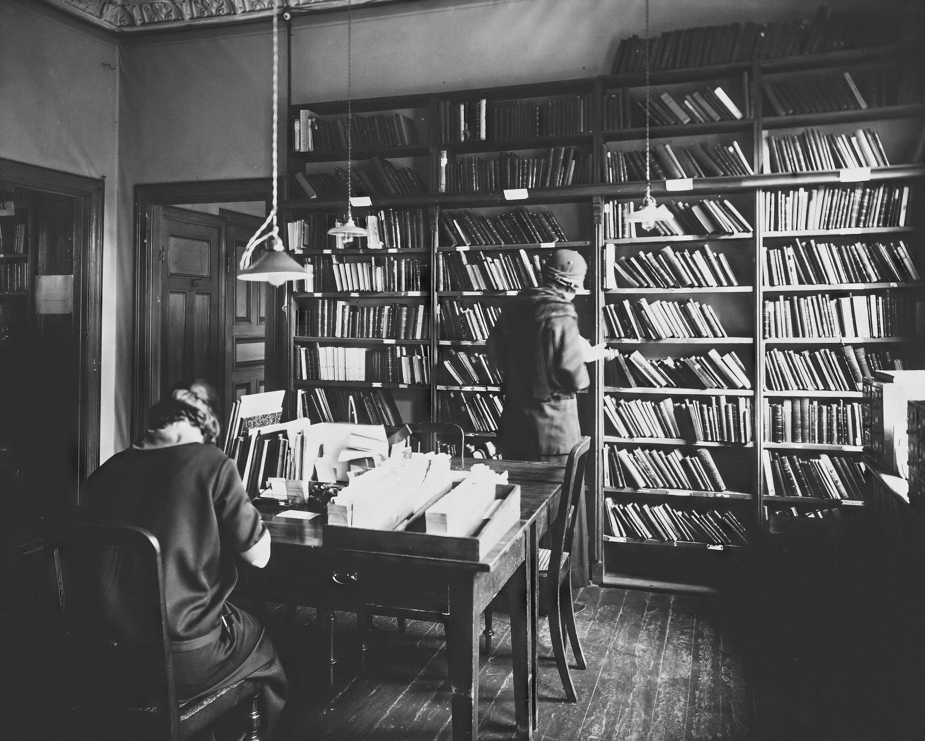 Two individuals immersed in a vintage library, one seated at a desk and the other browsing shelves filled with books. The ambiance reflects a timeless pursuit of knowledge.