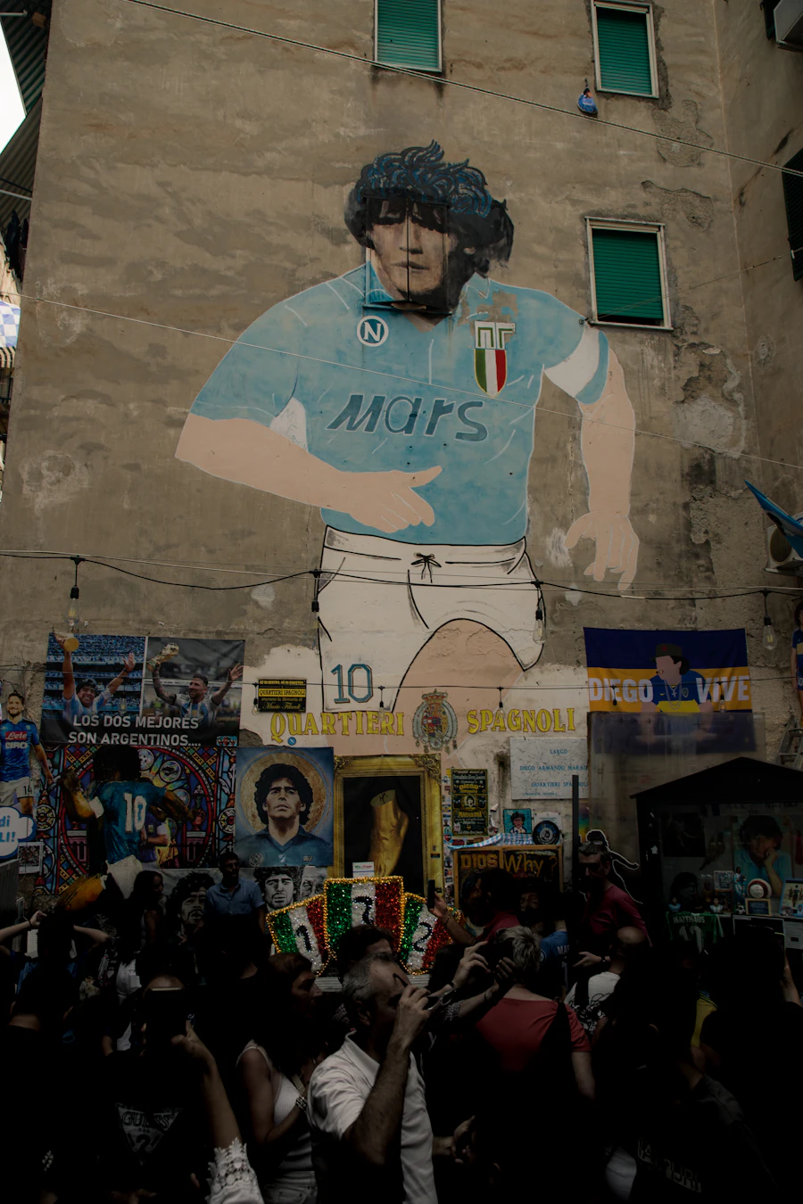 Large mural of Diego Maradona painted on a weathered building above a crowd of fans, with soccer posters and banners lining the lower wall.