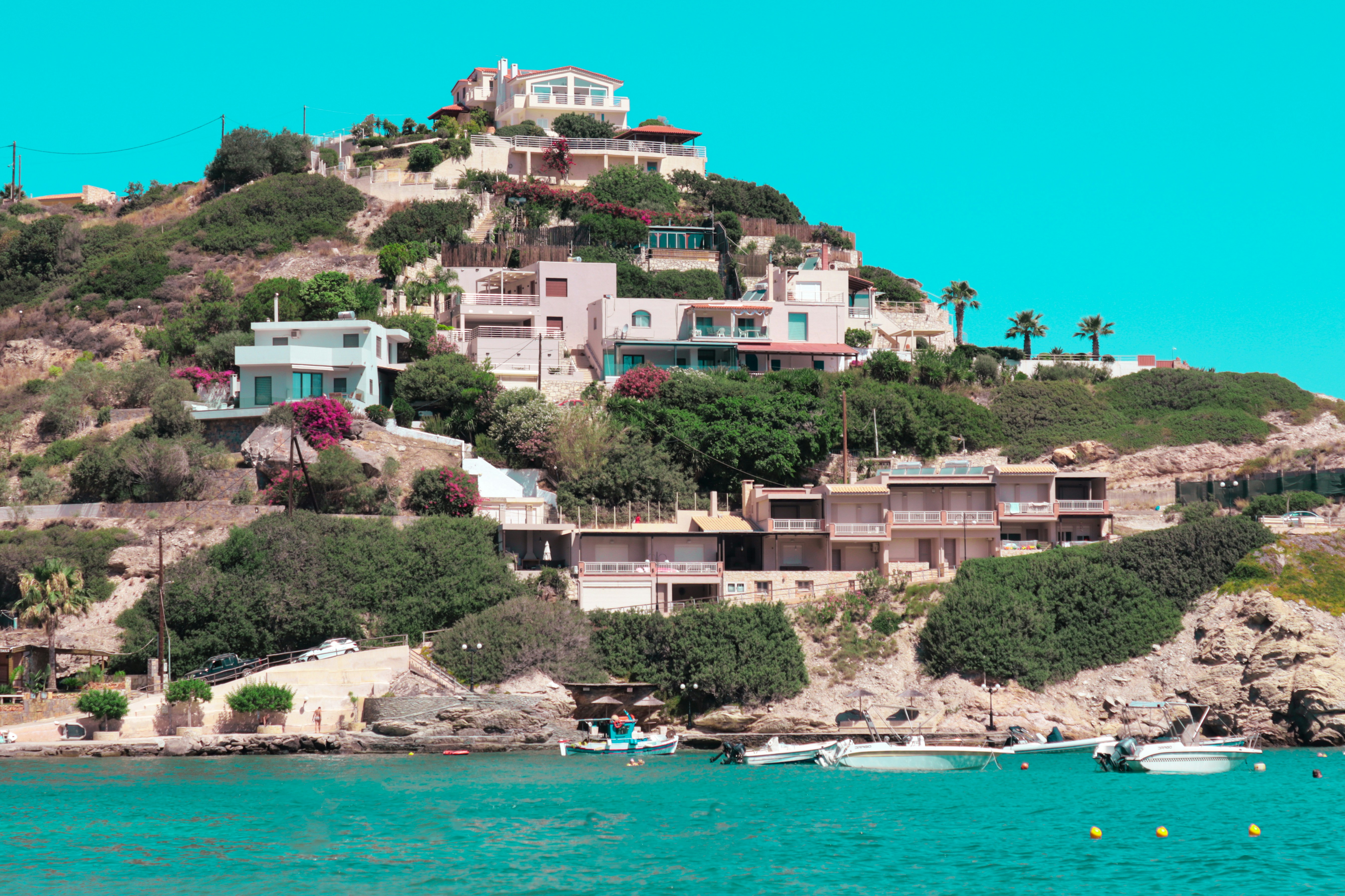 Colorful coastal homes nestled on a hillside, overlooking a tranquil turquoise sea. Lush greenery and palm trees enhance the picturesque scene.