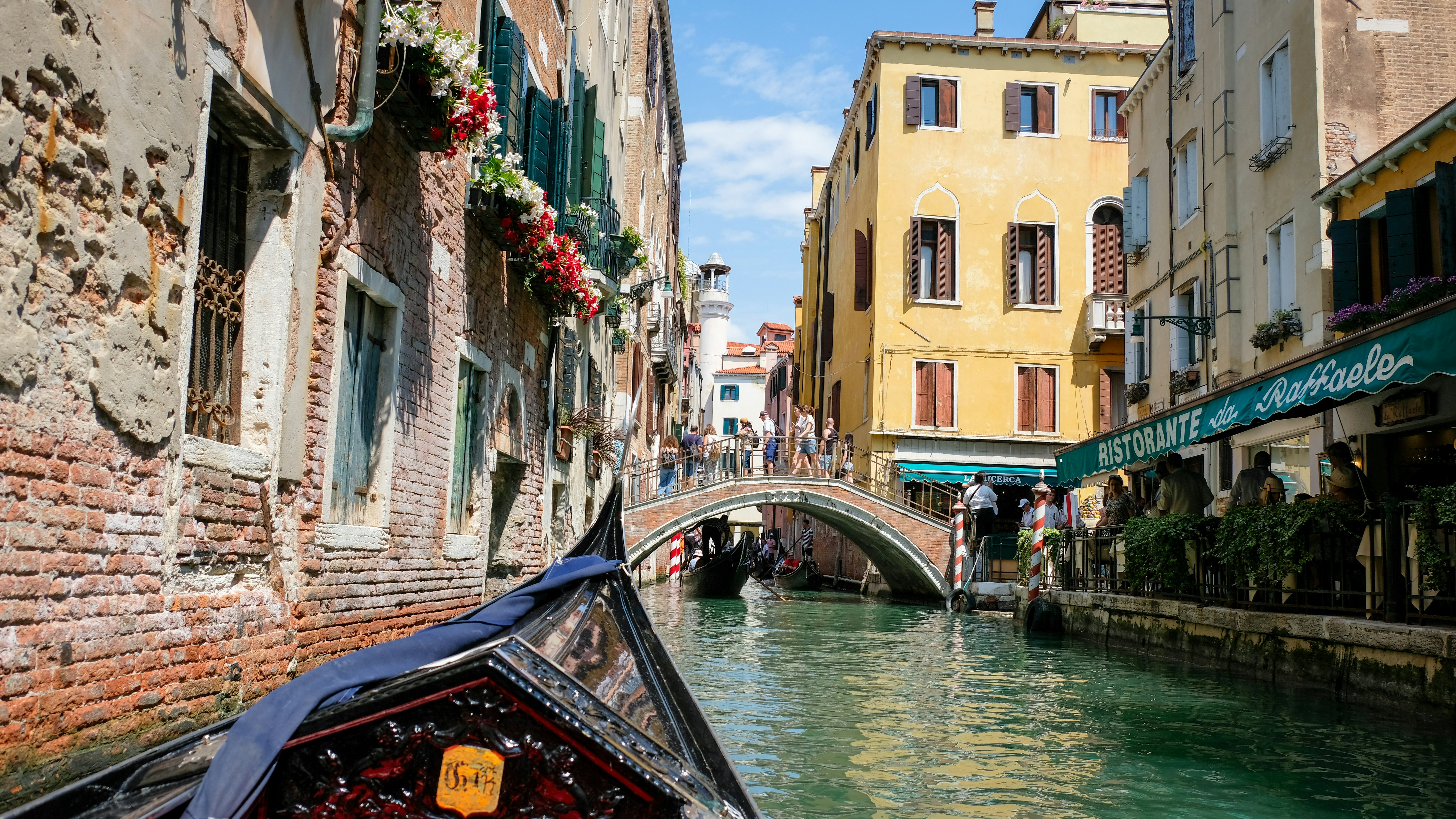 Gondola gliding through a narrow Venetian canal, flanked by charming buildings adorned with flowers. The scene captures the essence of everyday life in Venice.