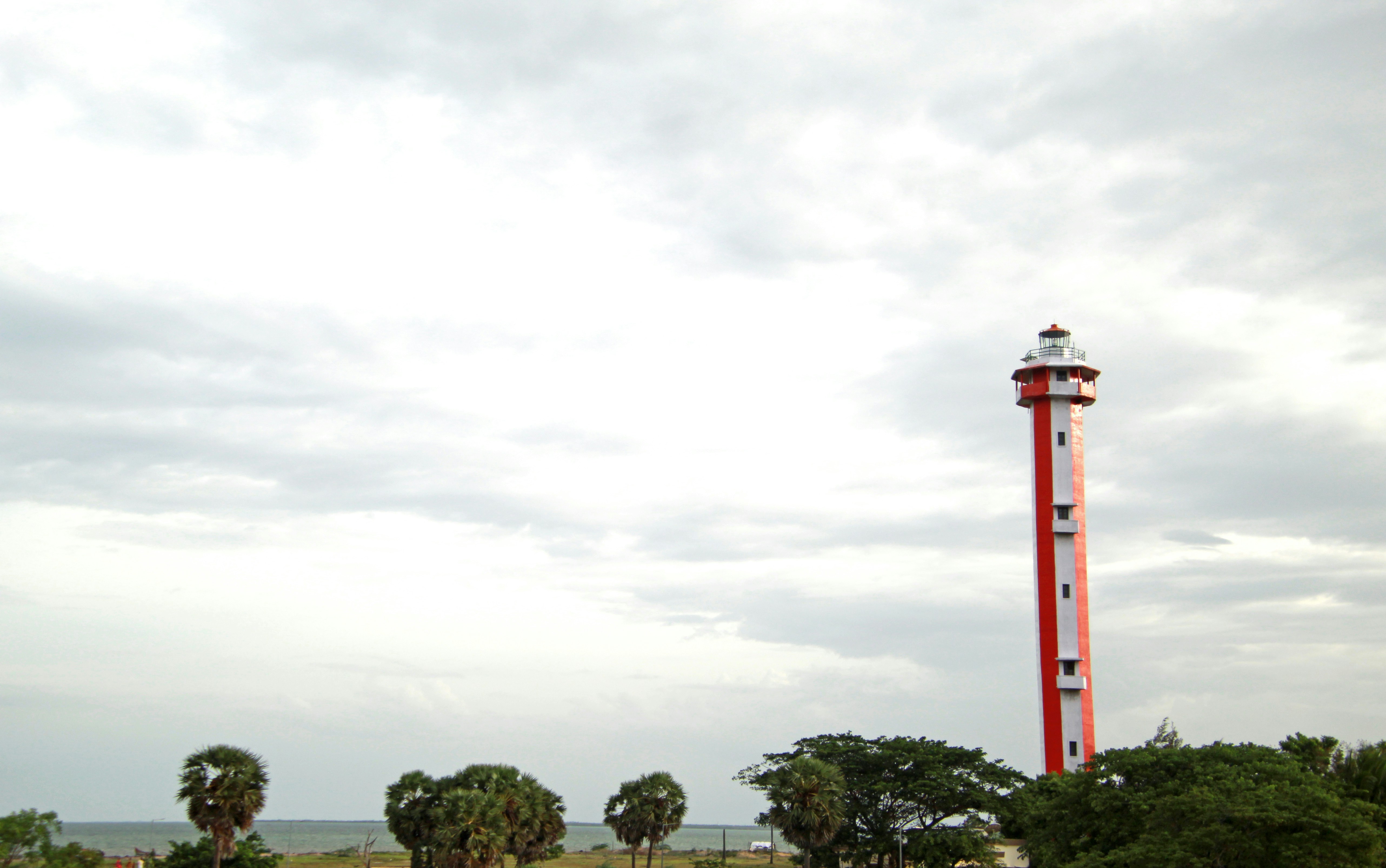 Crimson Beacon: Lighthouse Rising Over Palm Trees Under a Cloudy Sky