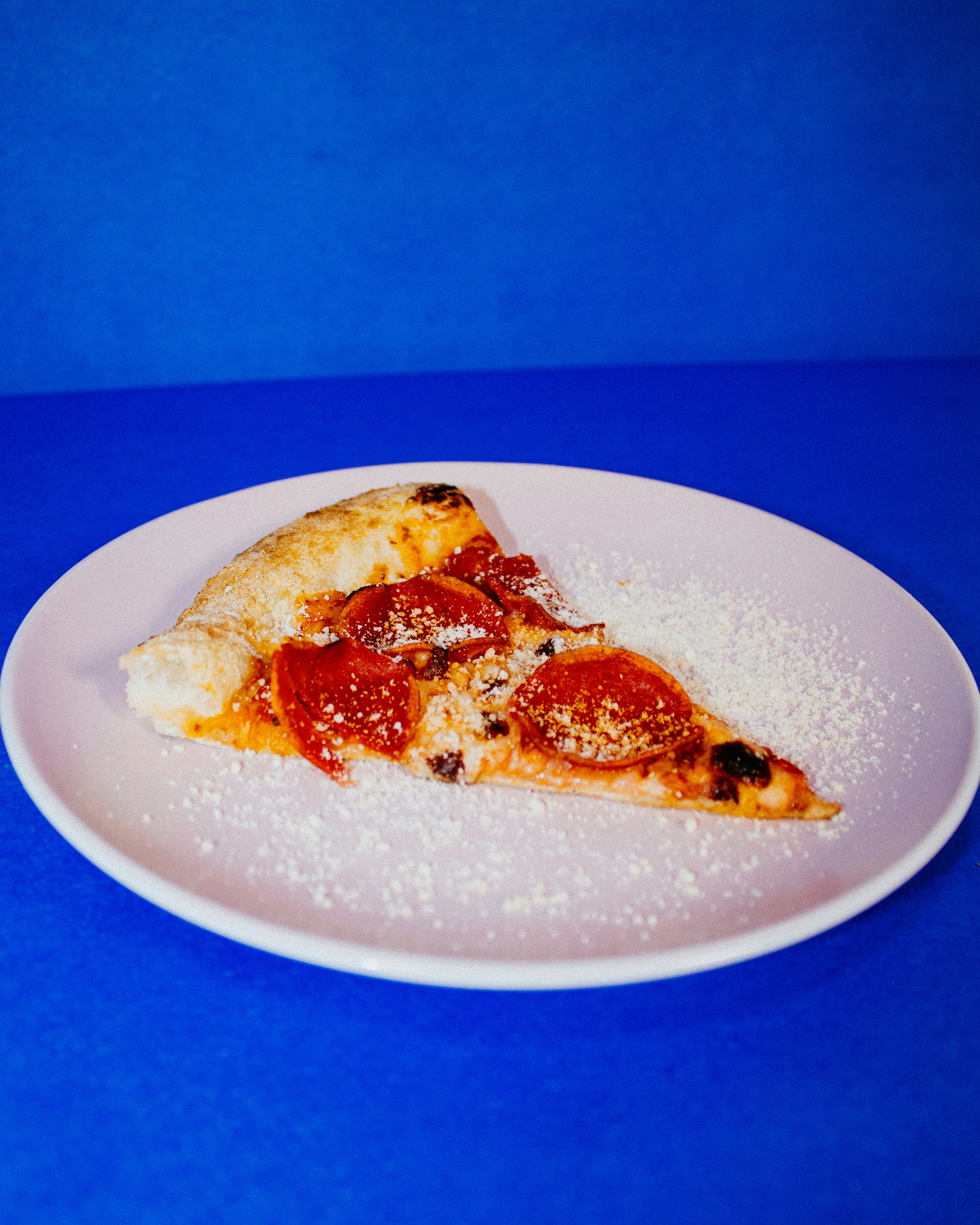 Single slice of pepperoni pizza resting on a white plate against a cobalt blue backdrop