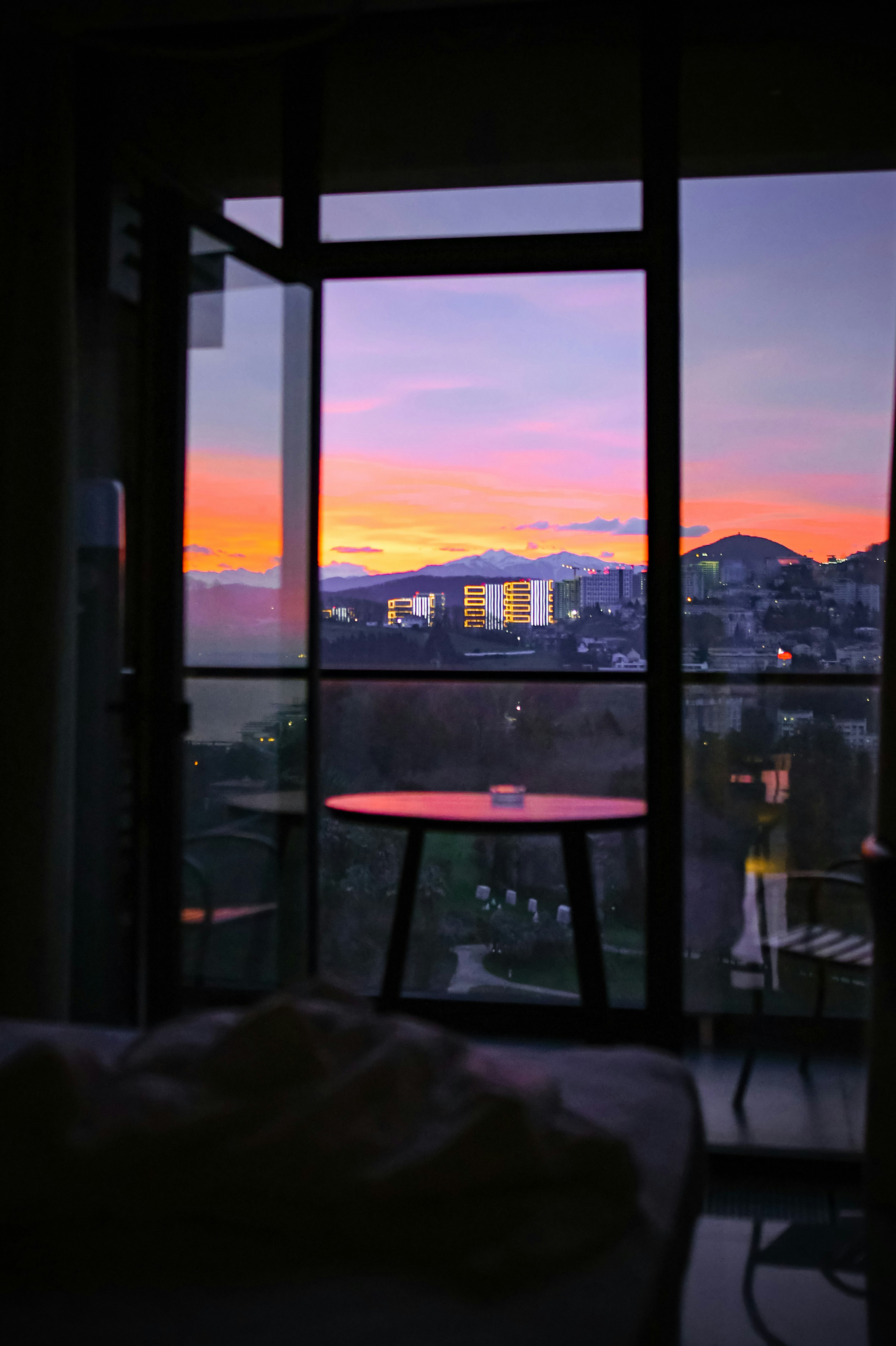 Sunset hues spill across a glass-walled interior, with a round pink table on the balcony foreground and distant city buildings beyond.
