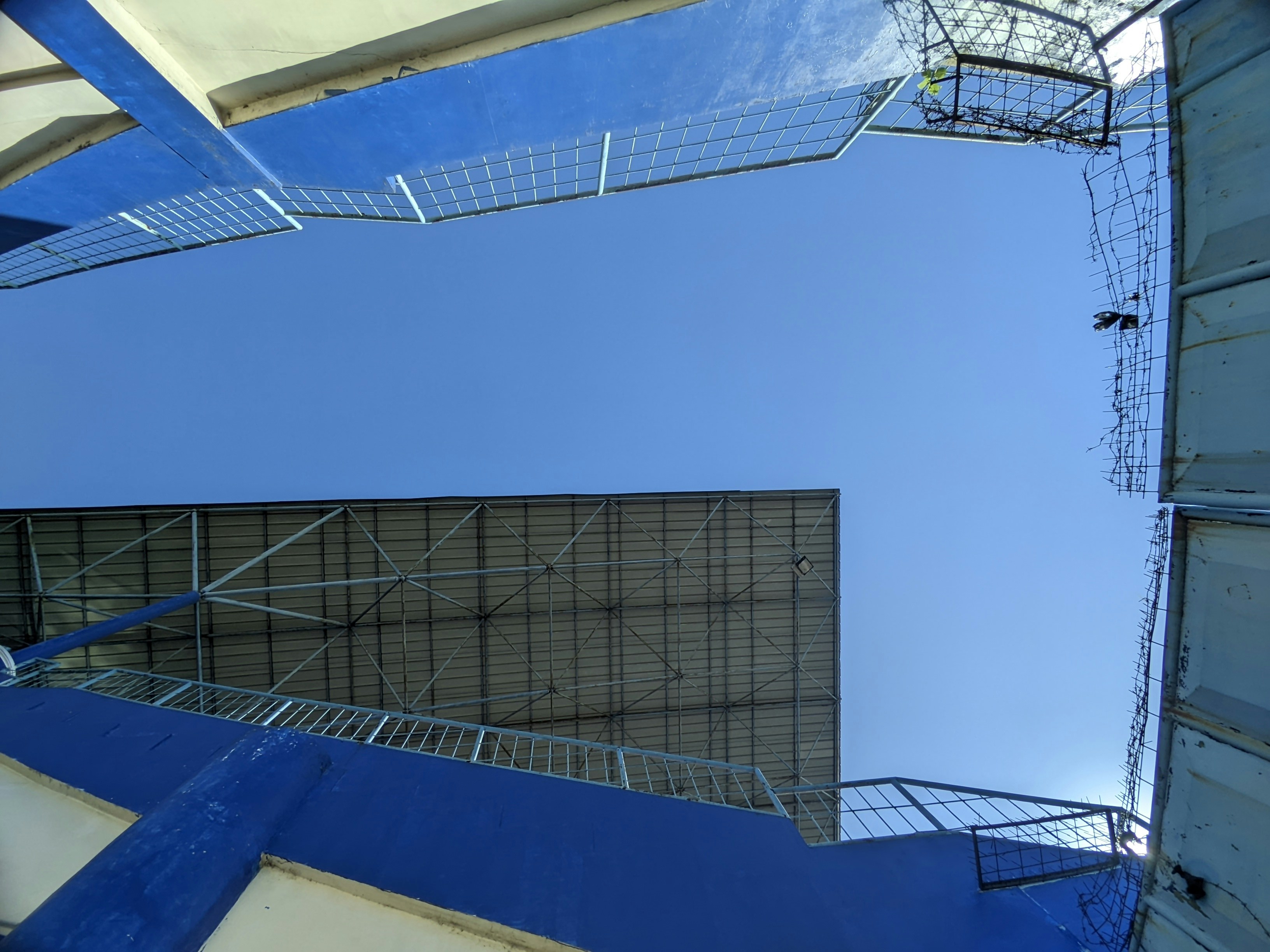 Blue-tinted urban canyon with tall buildings and metal railings framing a clear sky between them.