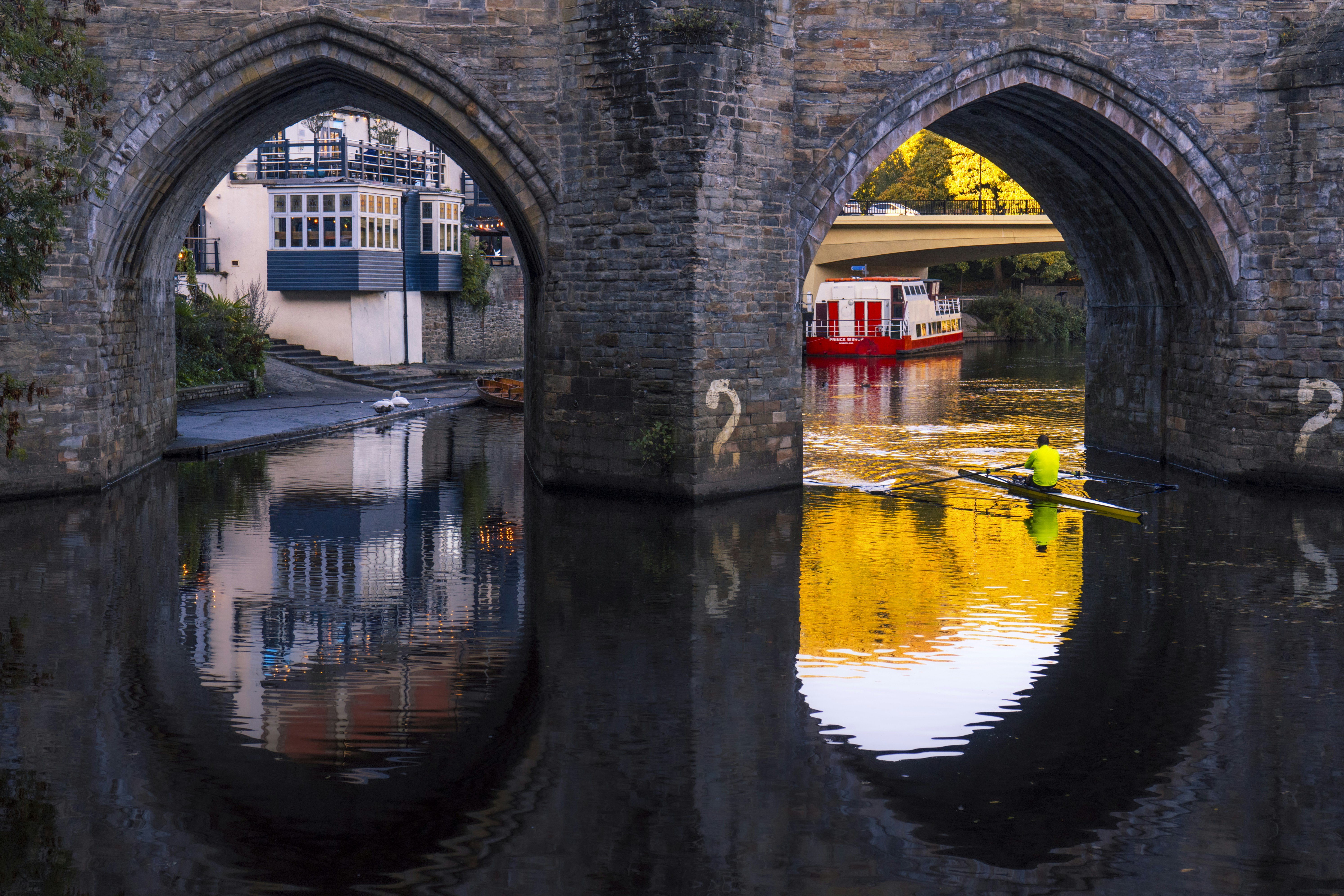 Rowboat gliding under a stone arch bridge, with vibrant reflections of buildings and autumn foliage mirrored in the calm water.