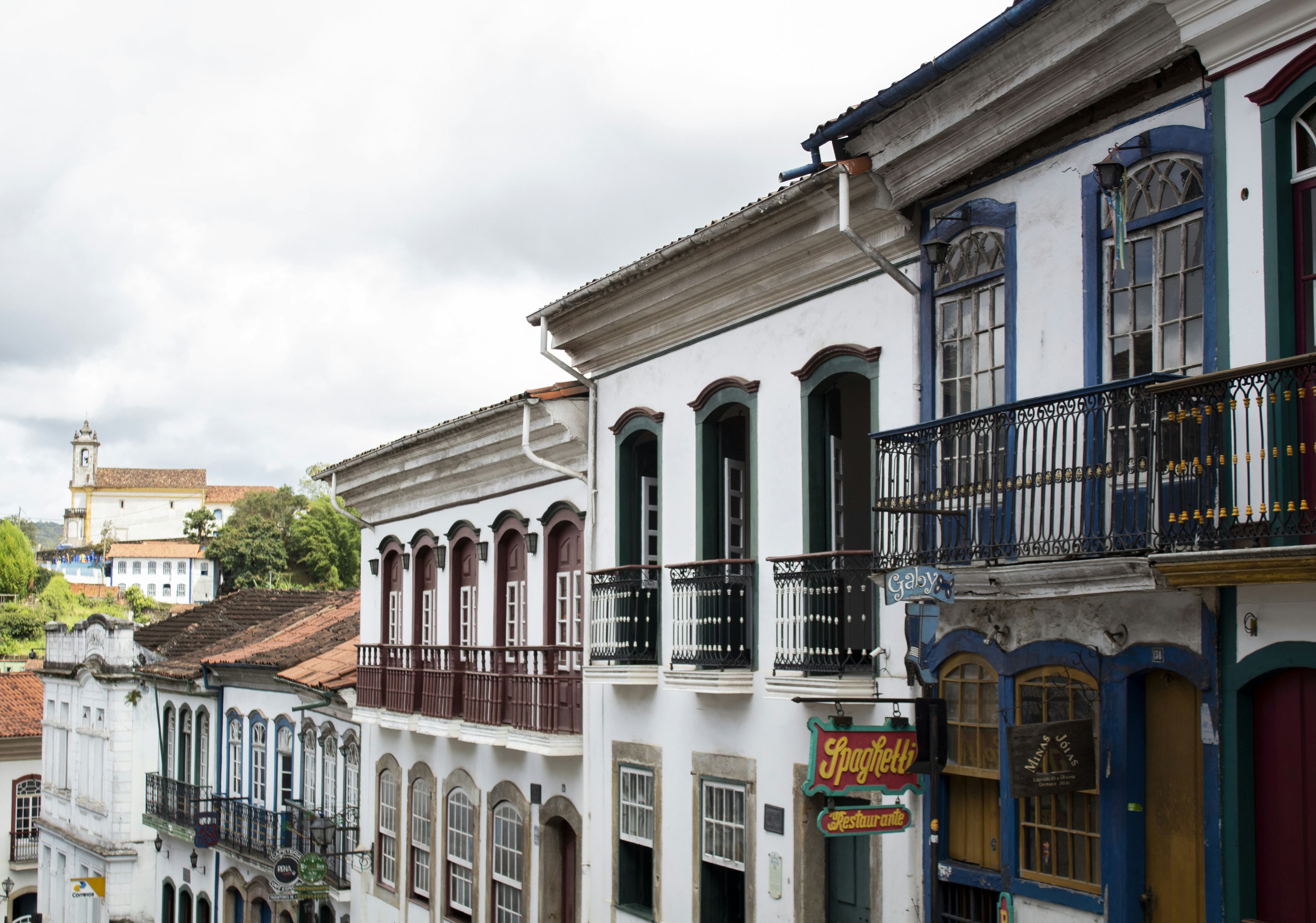 Colorful colonial buildings with ornate balconies line a sloping street under a cloudy sky.