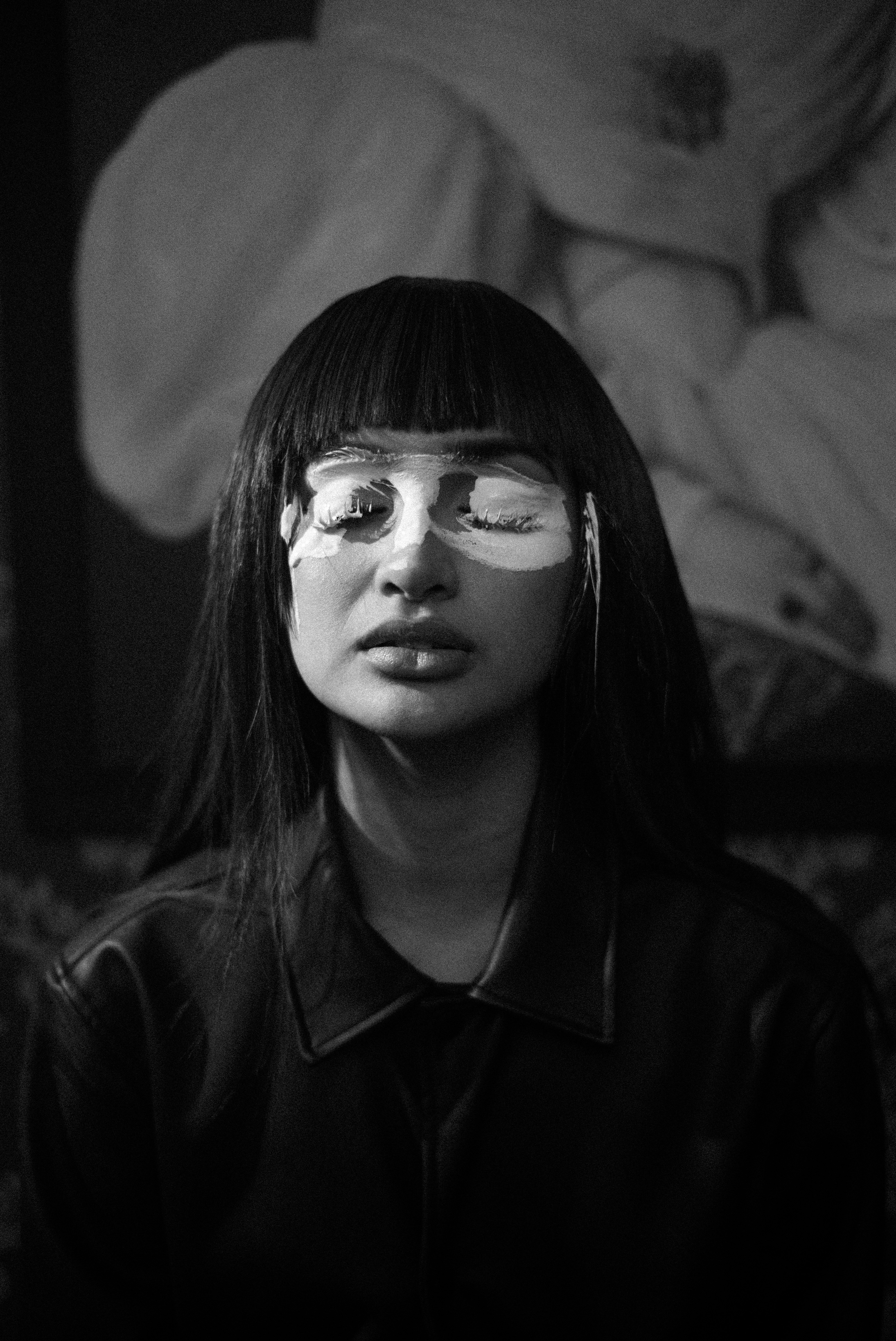 Black and white portrait of a woman with painted eyelids and straight bangs, set against a blurred background.