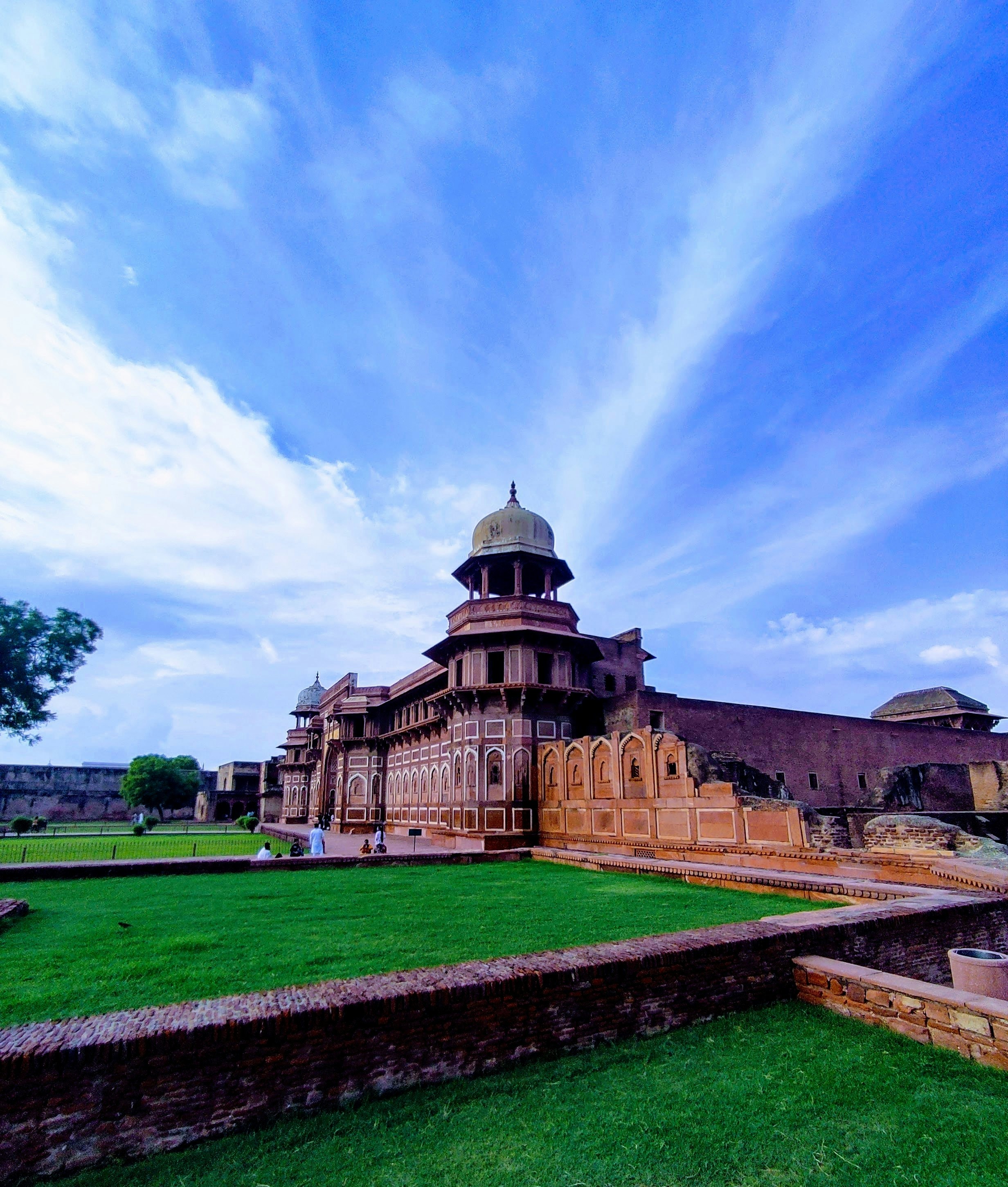 Historic sandstone fortress with a central domed pavilion rises above a manicured lawn under a vivid blue sky.