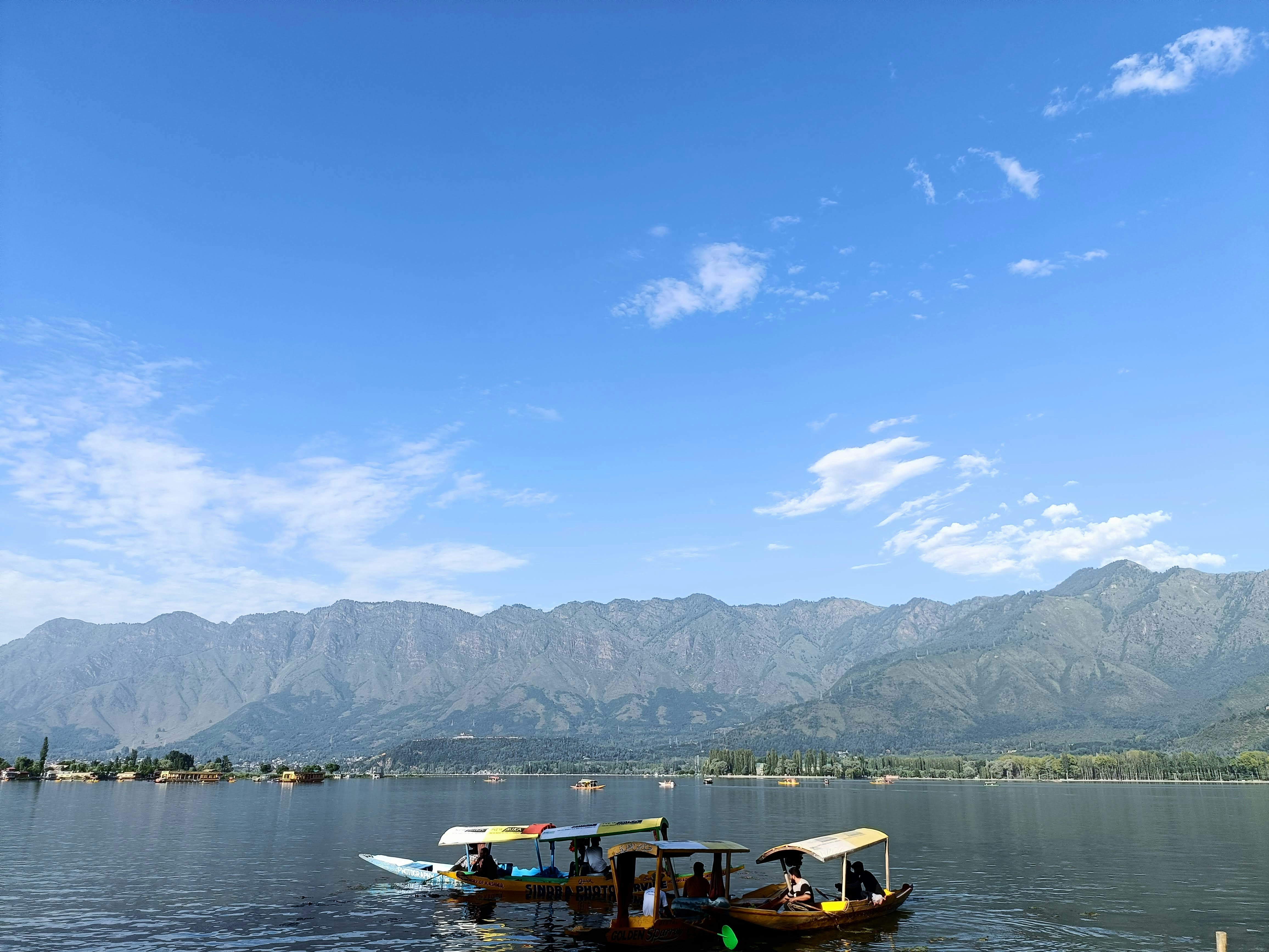 Traditional wooden shikaras gliding on the calm waters of Dal Lake, framed by majestic mountains and a clear blue sky.