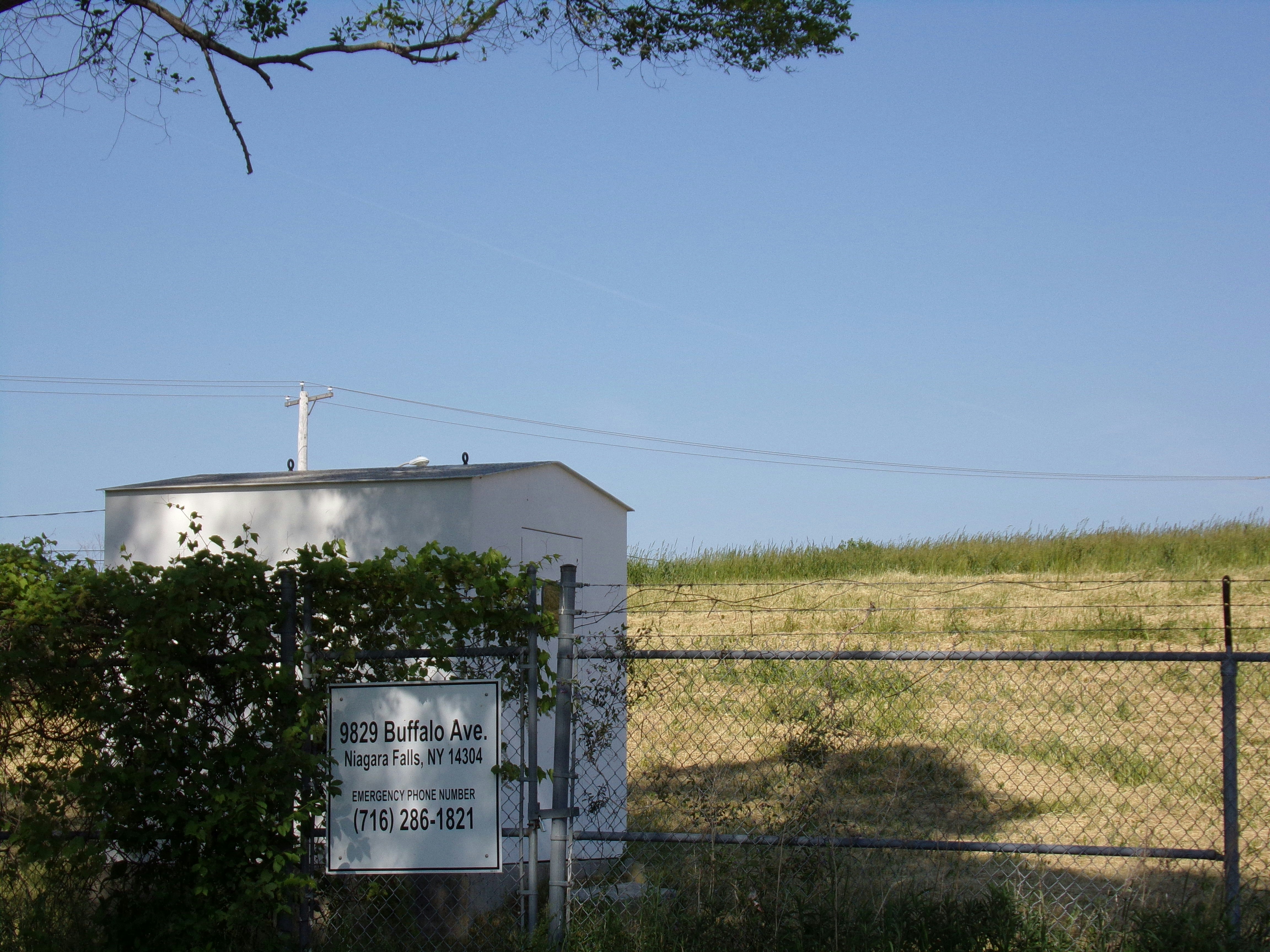 Photograph shows a weathered utility box behind a chain-link fence, with a grassy slope and a clear blue sky.