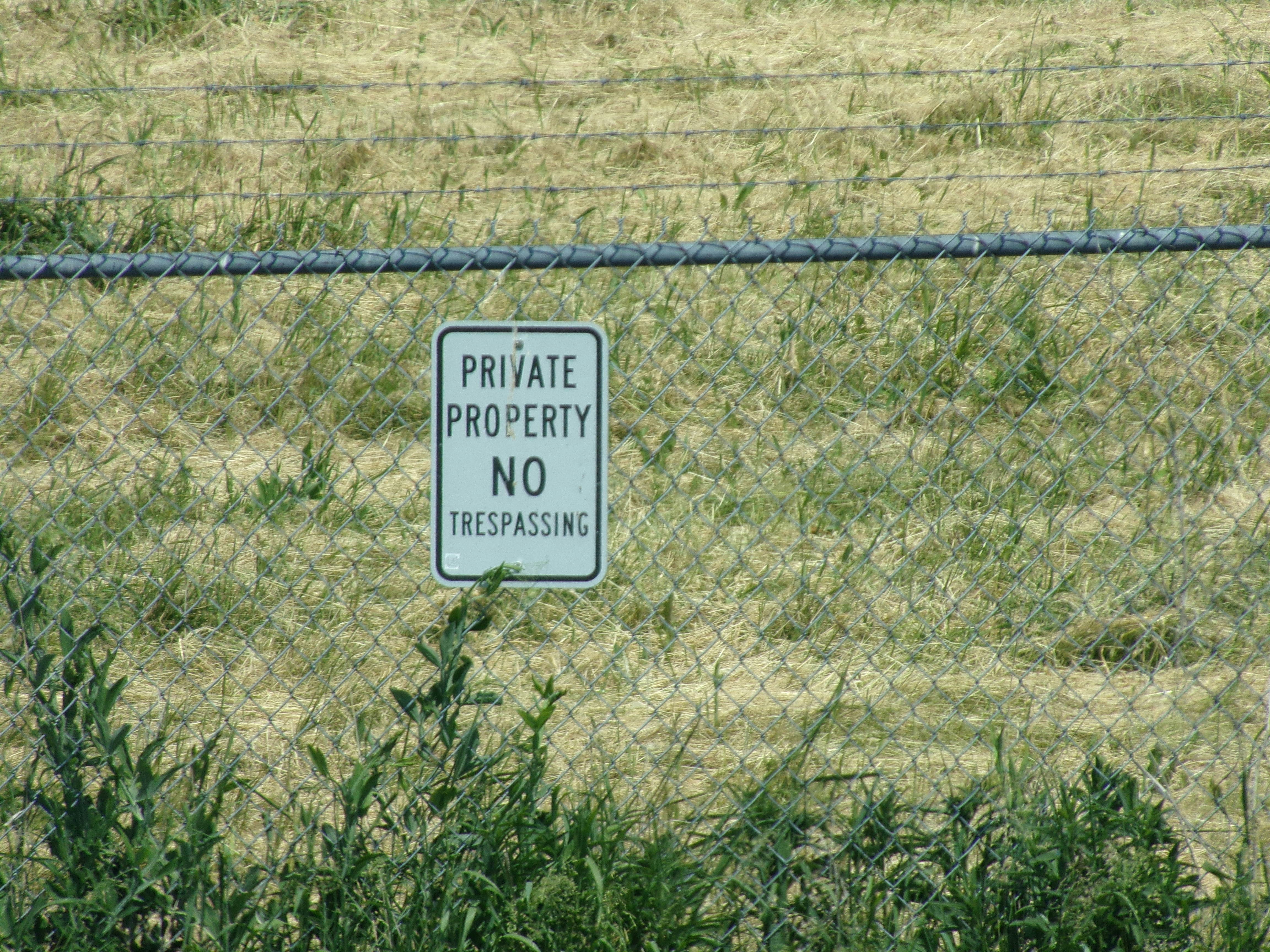 Sign indicating private property and no trespassing, surrounded by grass and a chain-link fence.