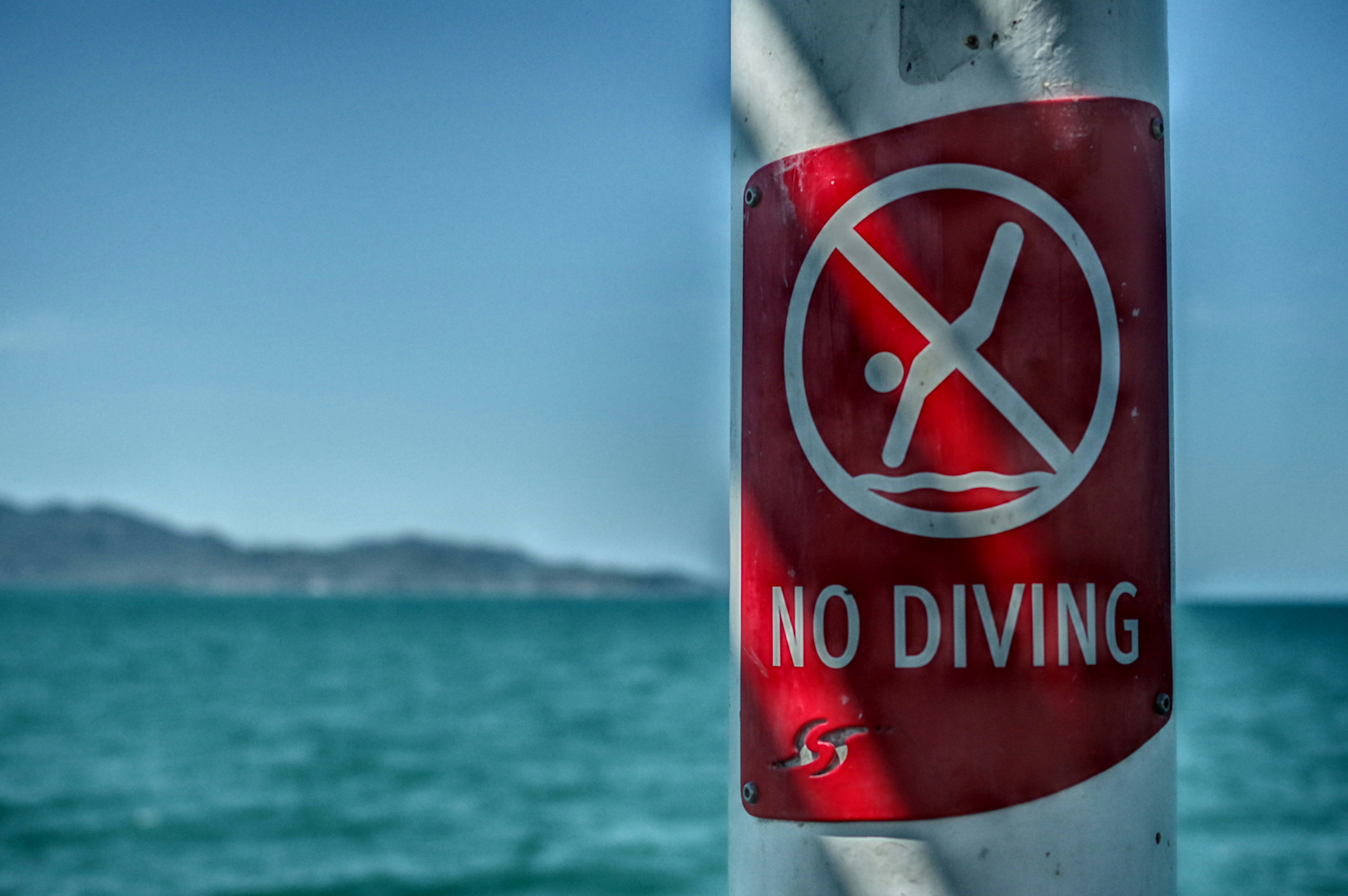 Red and white 'No Diving' sign on a pole near a turquoise sea with distant mountains.