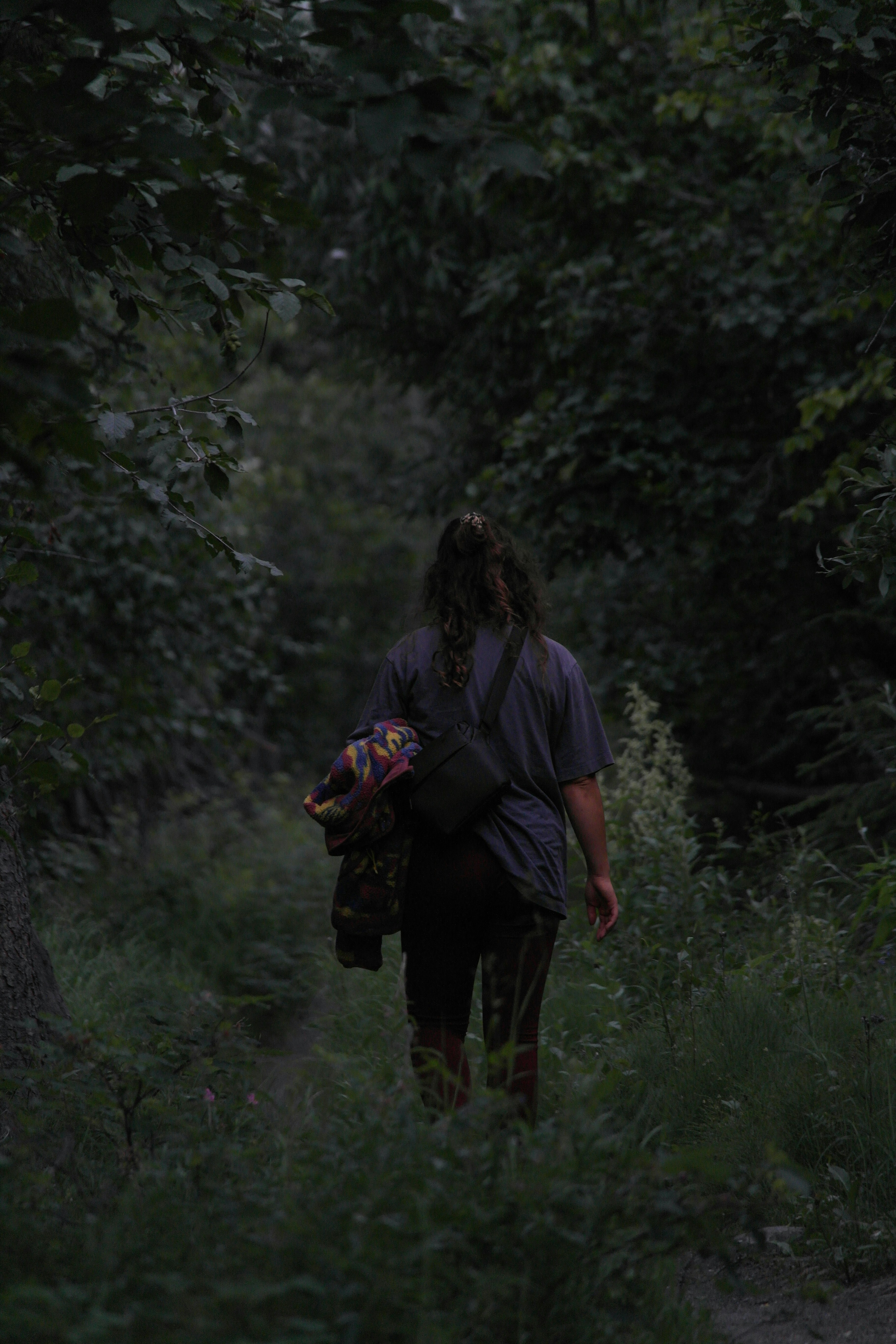 A person seen from behind walks along a narrow, overgrown forest path at dusk, carrying a bag and a colorful jacket.