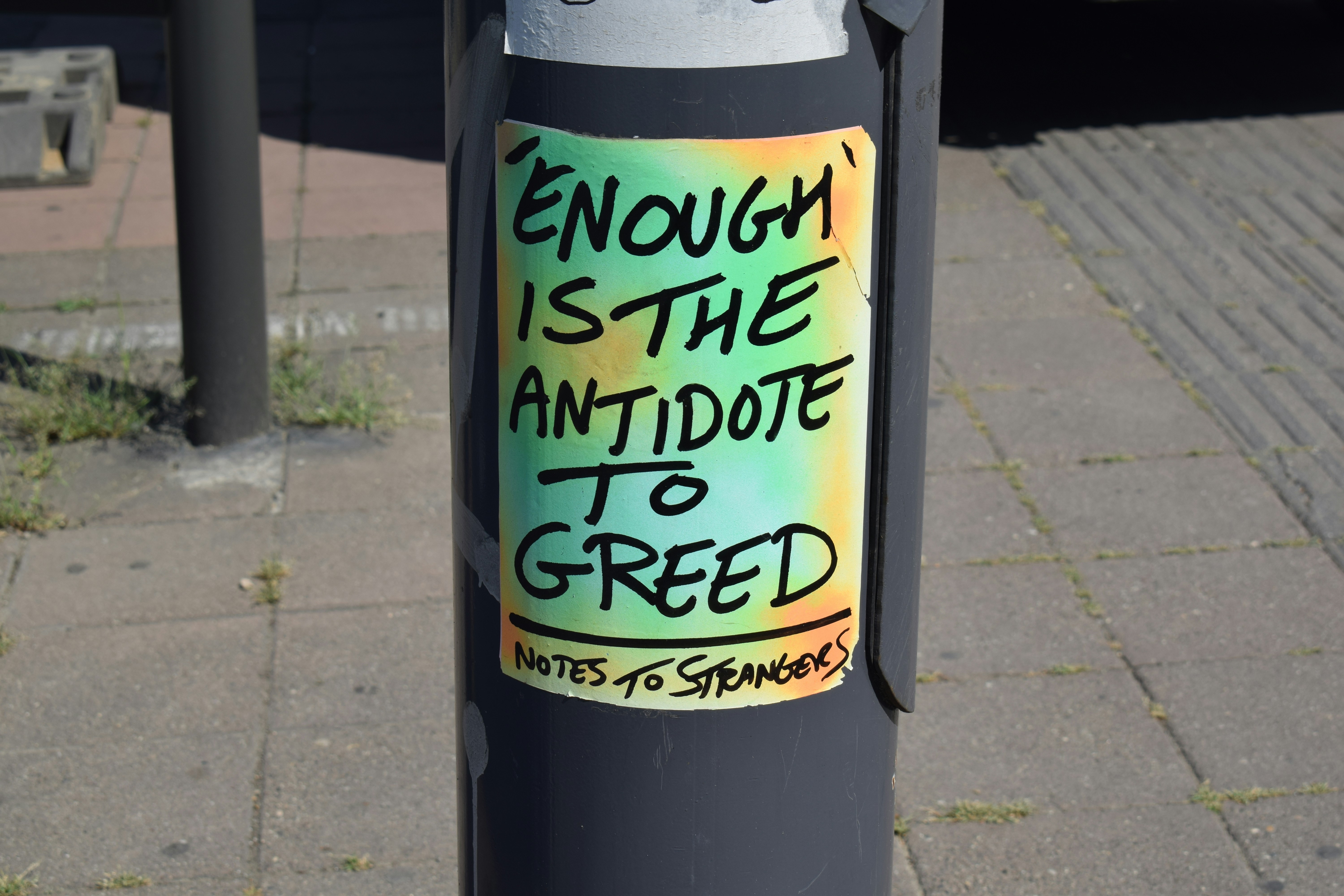 Colorful sticker proclaiming 'ENOUGH IS THE ANTIDOTE TO GREED' affixed to a utility pole. A thought-provoking message for passersby.