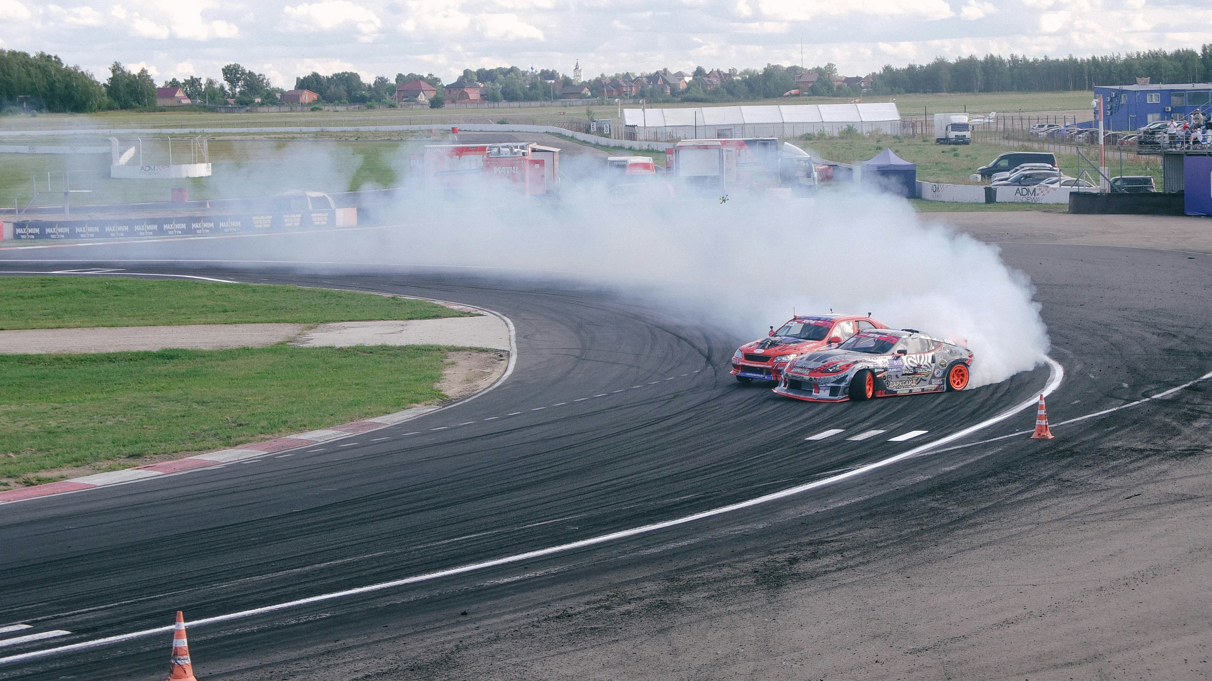 Red race car drifts through a tight turn on a race circuit, tires billowing white smoke as an orange cone marks the apex. A trailing car and distant grandstands complete the dynamic track backdrop.