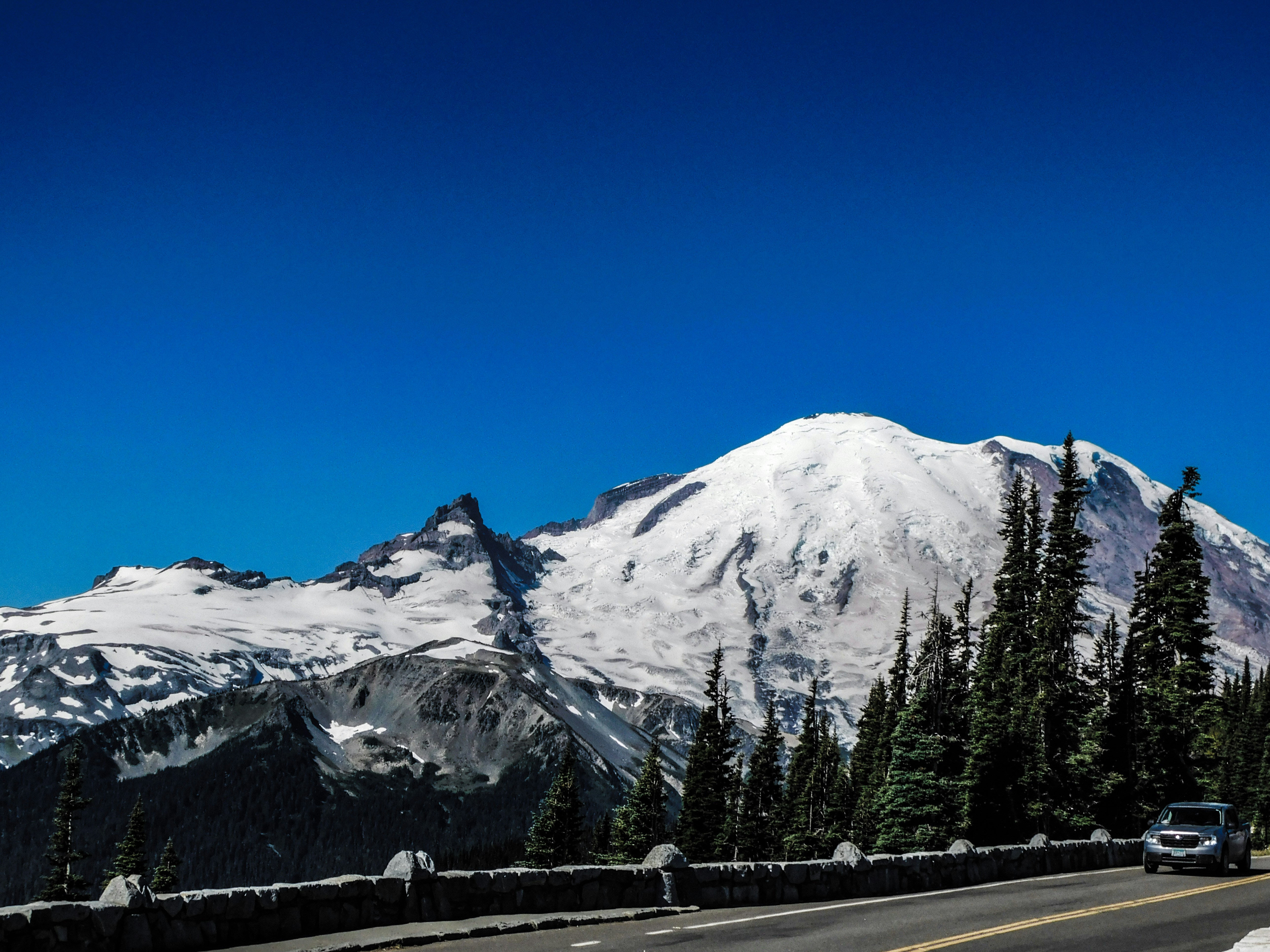 Snow-dusted mountain peak along a winding road with evergreen trees lining the foreground, under a clear blue sky.
