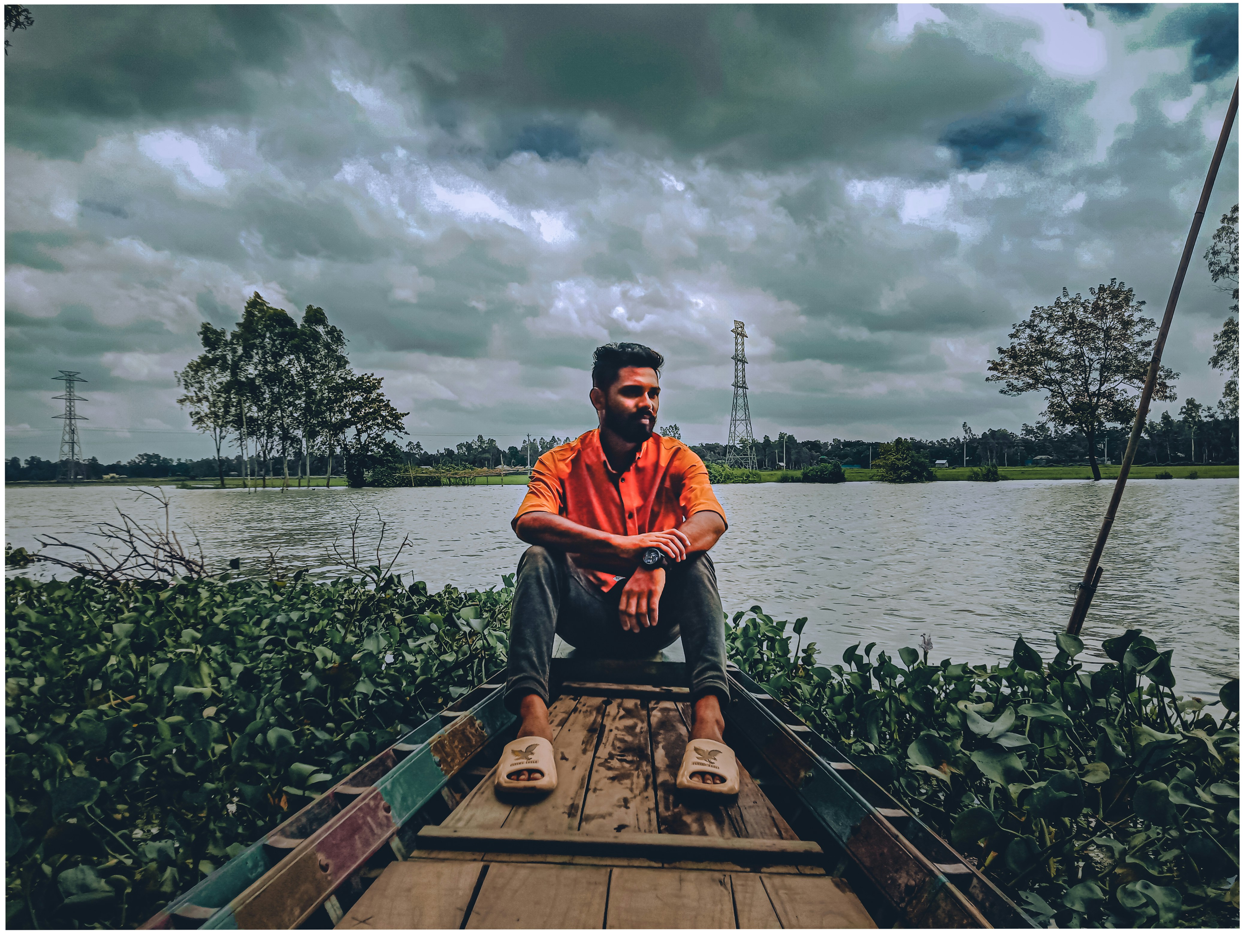 Man in an orange shirt sits on a boat amidst lush river scenery under dramatic clouds.