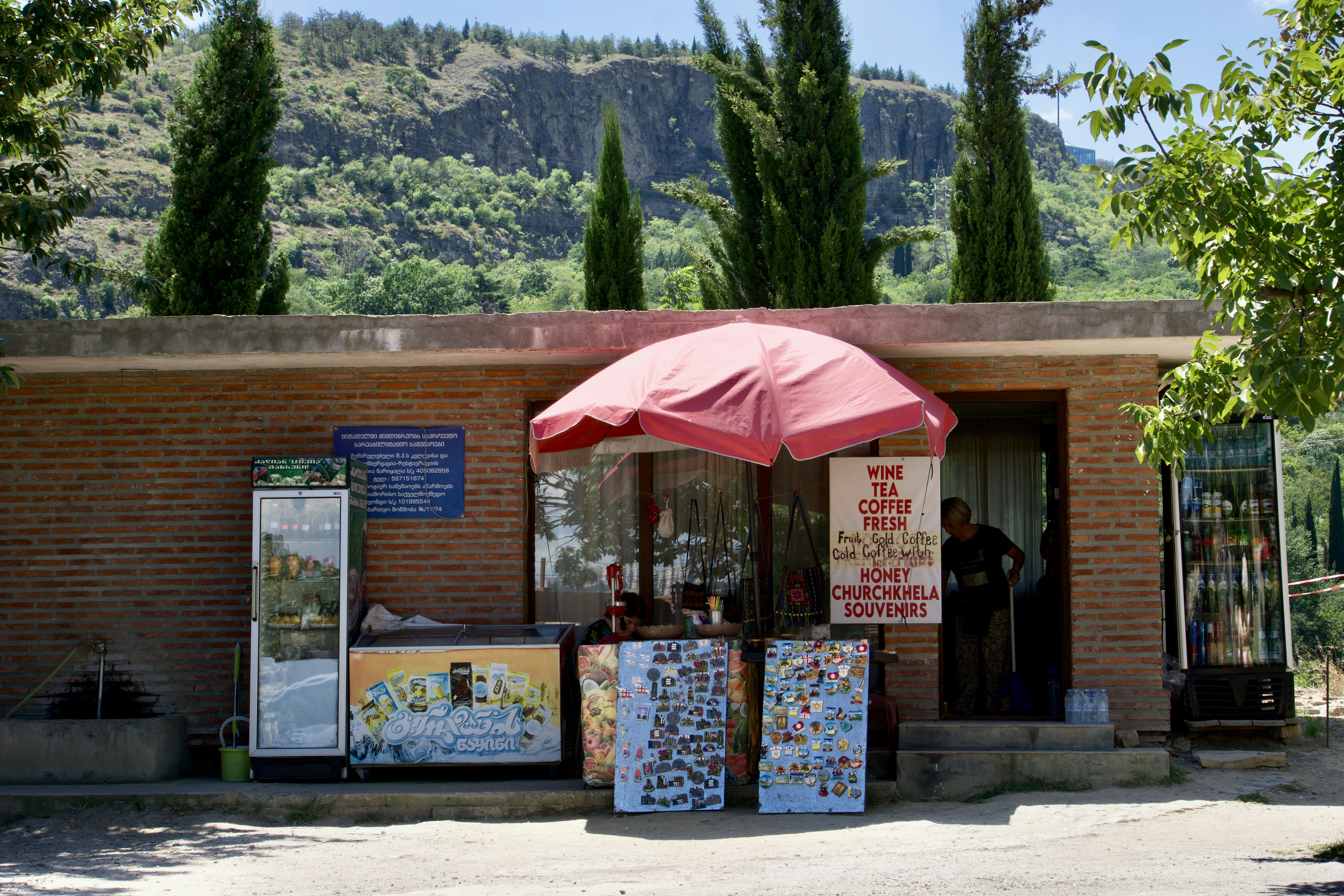 Small brick storefront with a red umbrella, surrounded by lush greenery and mountainous backdrop.