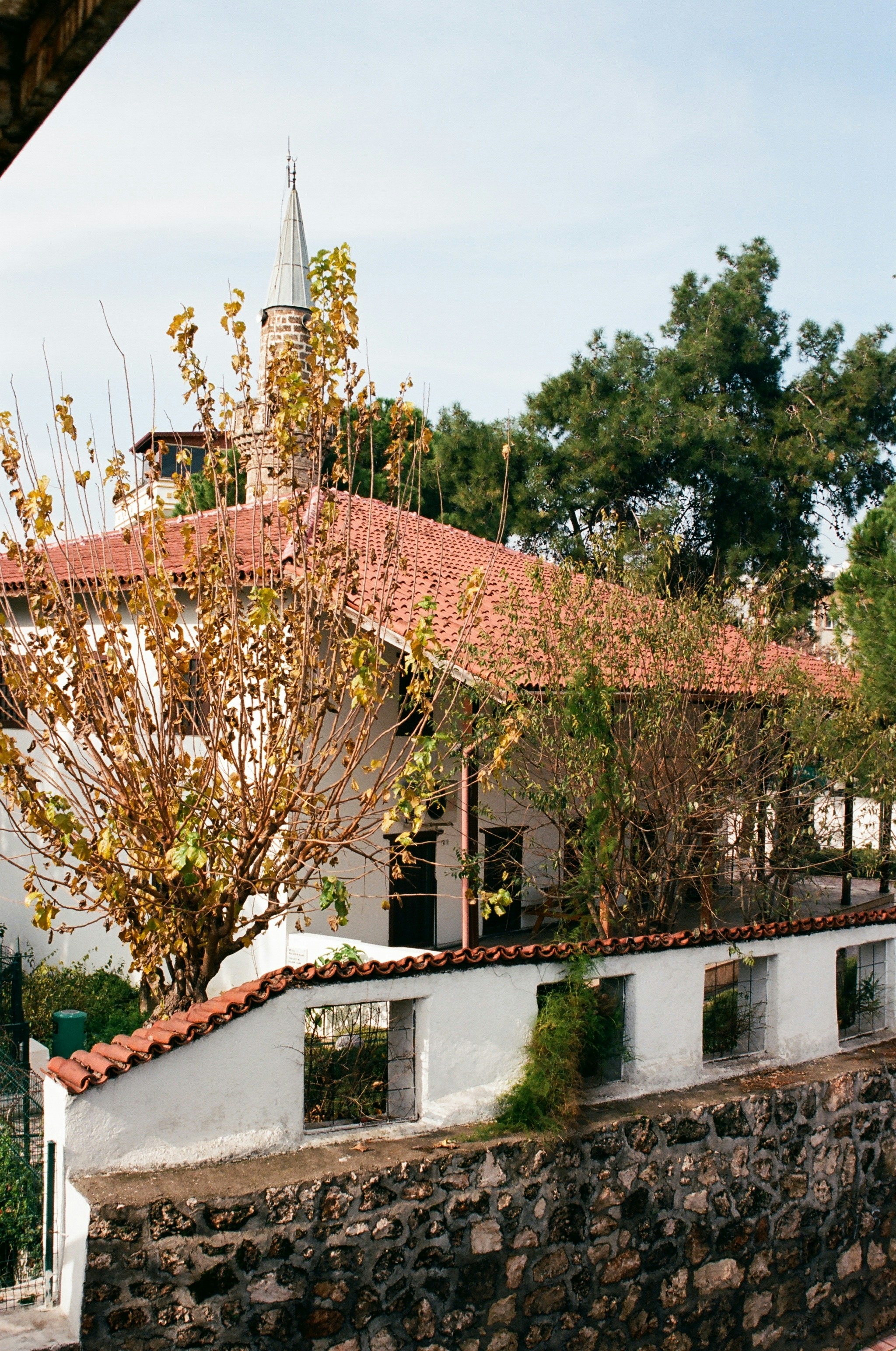 Photograph of a white-walled building with a red-tiled roof and a slender church spire rising behind autumn trees. The shot is framed by a stone wall in the foreground, capturing a quiet village edge bathed in soft sunlight.