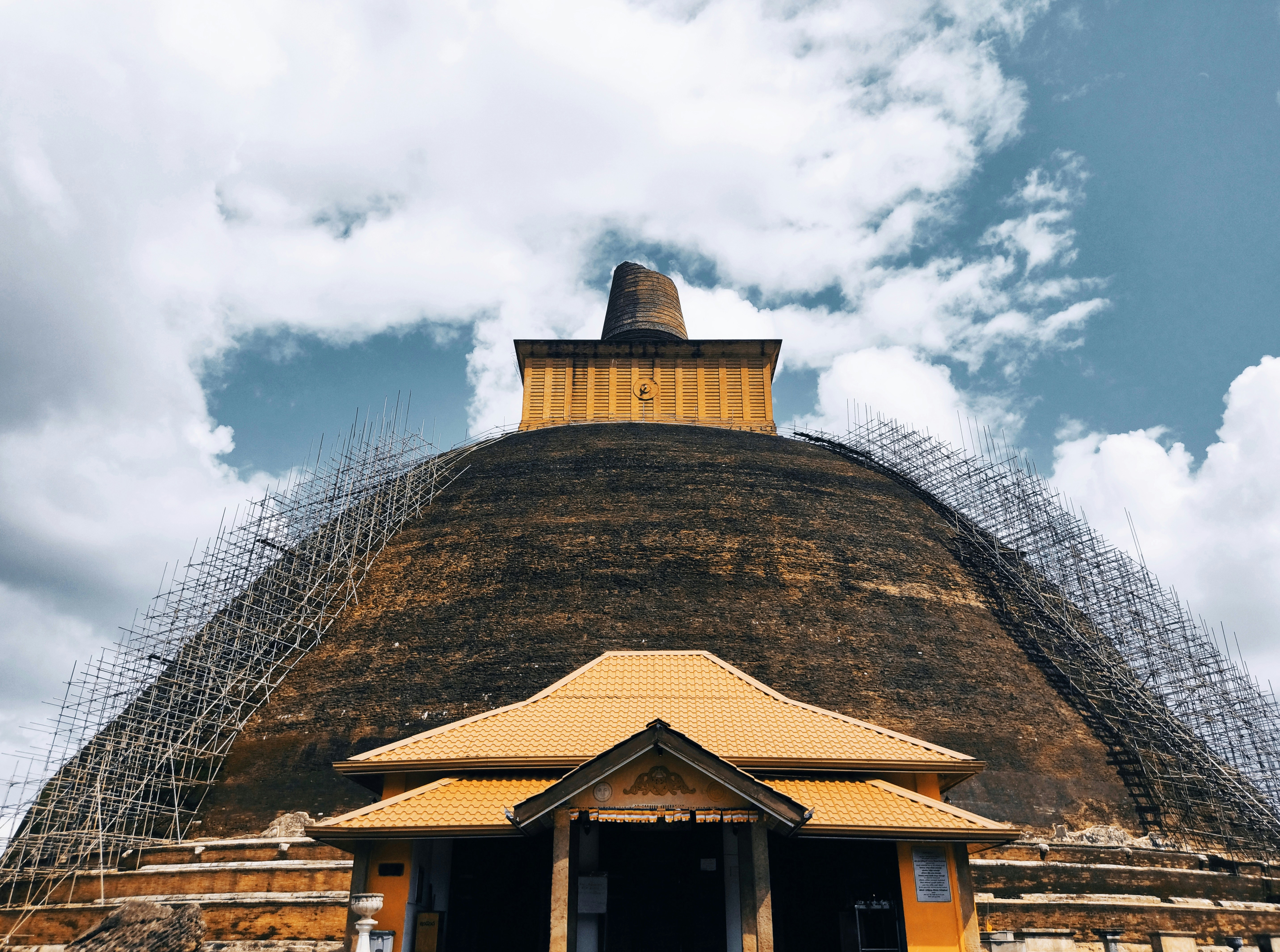 Photograph of a large, domed stupa surrounded by scaffolding. The structure sits at the base with a small entrance, set against a bright blue sky with clouds.