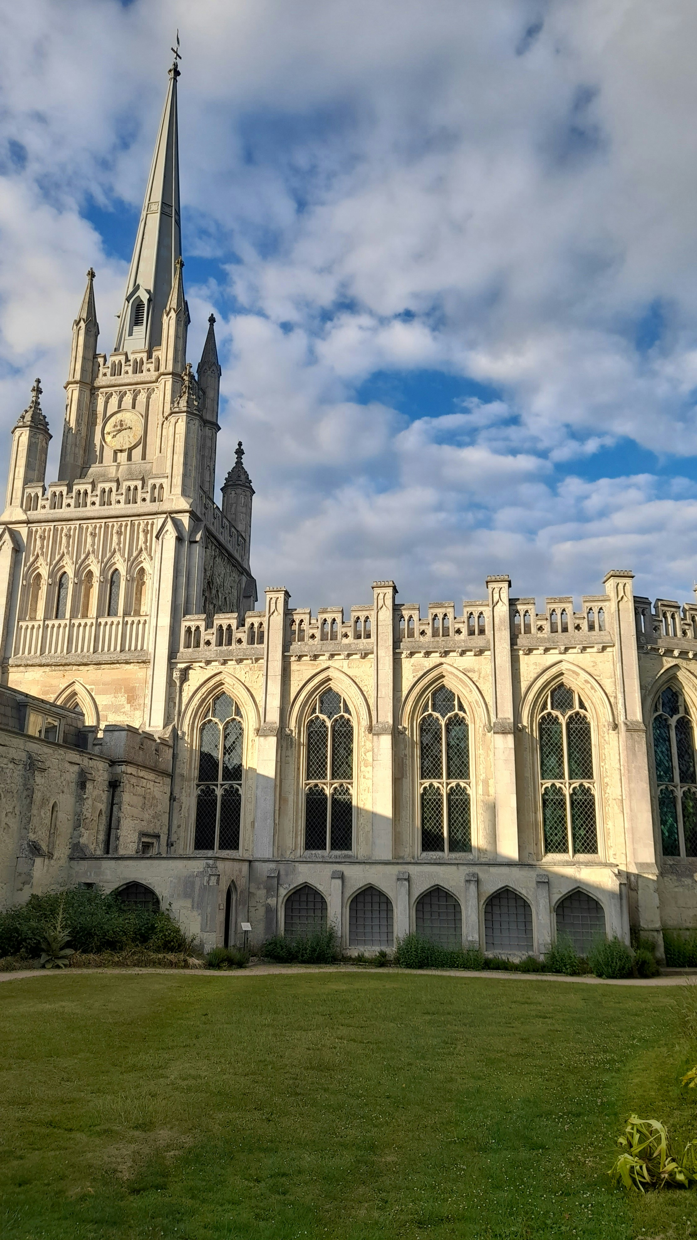 Gothic cathedral facade with a tall spire and pointed arches dominates the left side, set against a bright blue sky. A tidy lawn and cloister frame the historic stonework.