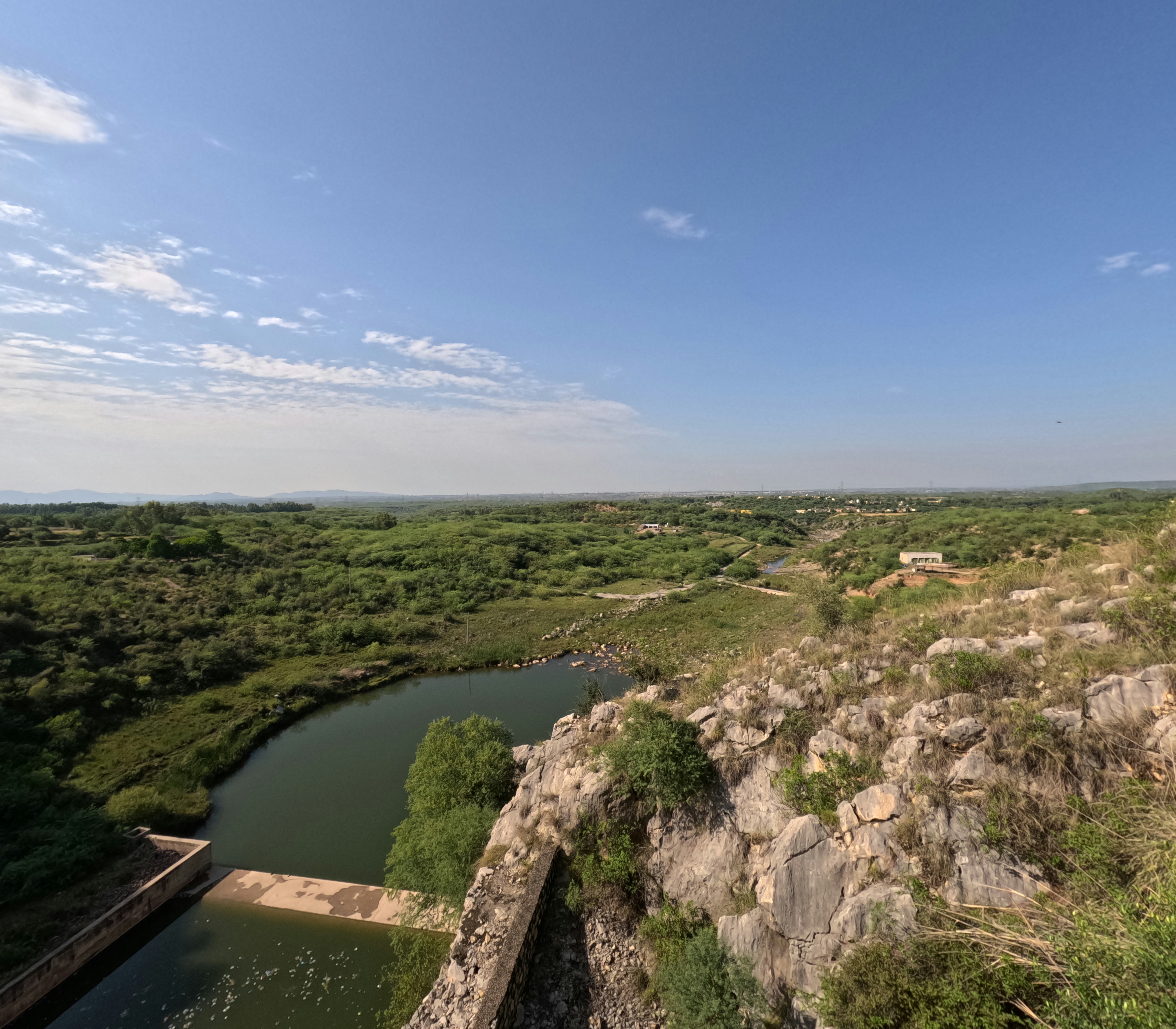 Sunlit rocky promontory overlooks a winding river through green scrub with distant fields, a serene landscape photograph.