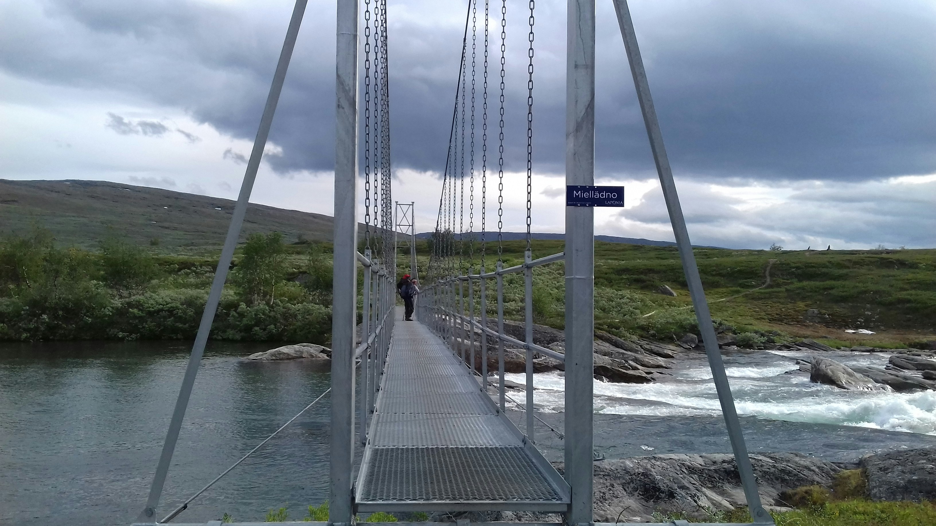 Steel suspension footbridge spans a shallow river through open moorland under a cloudy sky. A lone pedestrian walks toward the camera along the grated walkway.