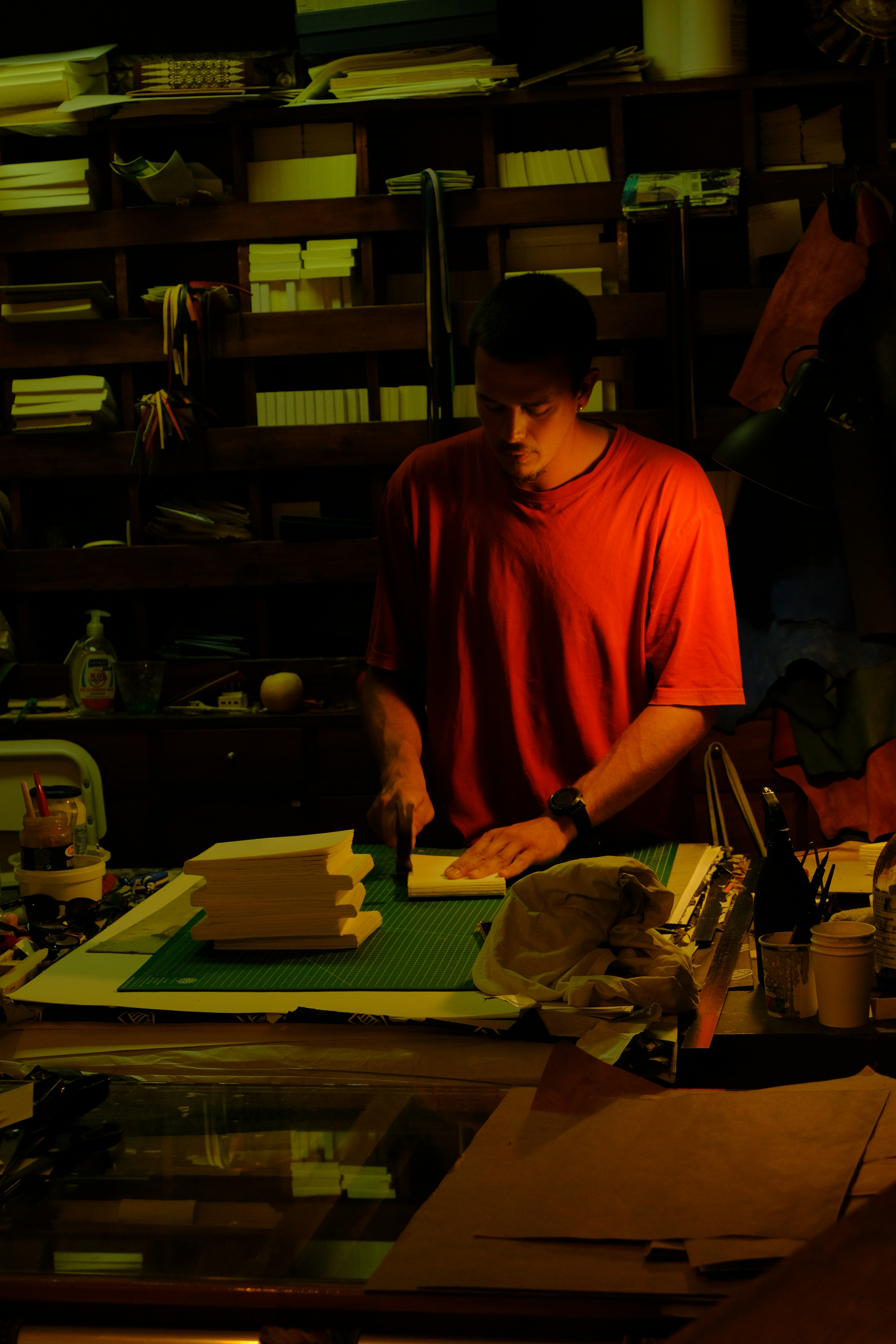 A man in an orange T-shirt works at a cluttered workshop desk, trimming stacked pages under warm amber lighting. Shelves laden with papers and tools form the busy background.
