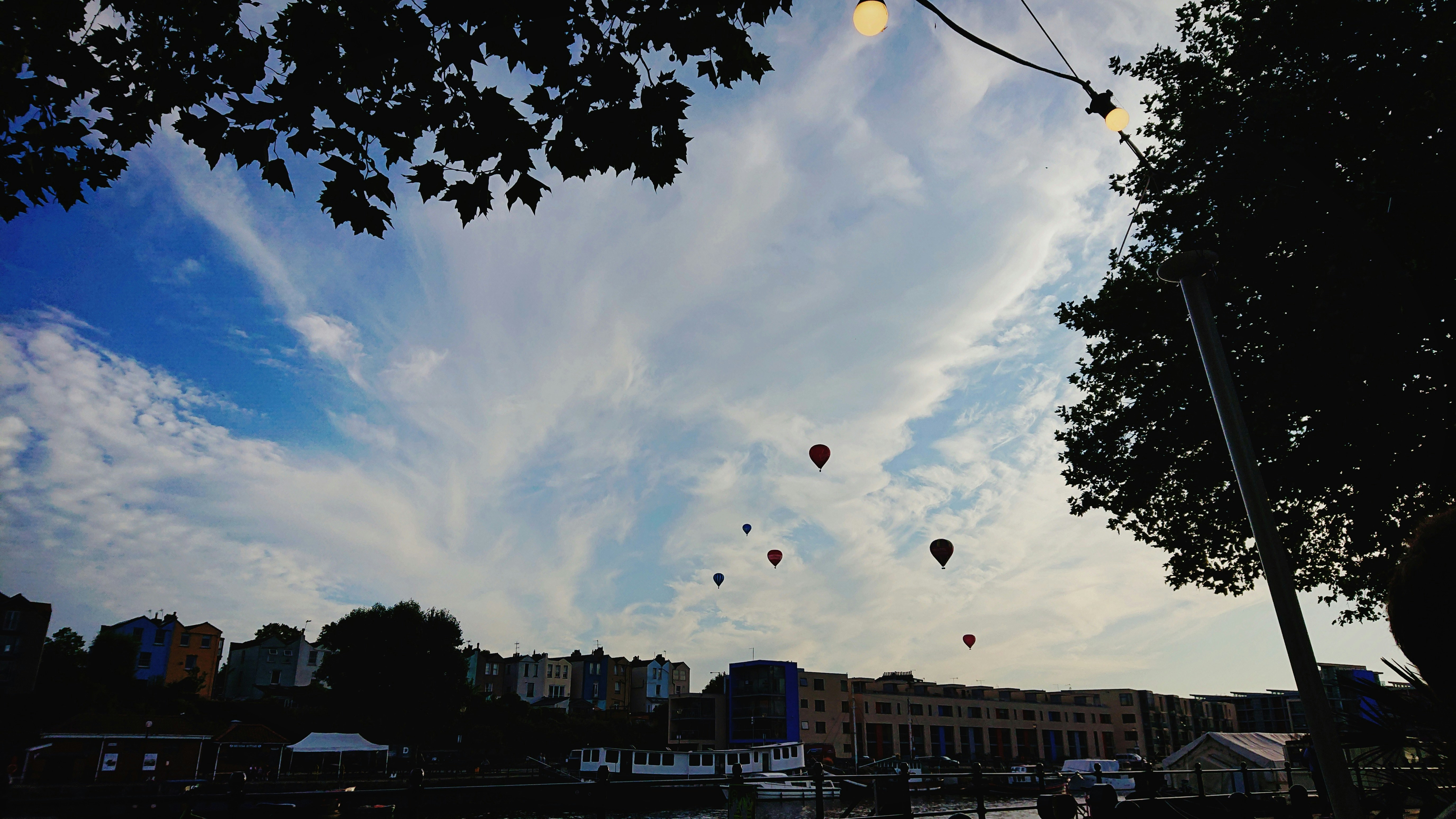 Colorful balloons drift over a riverside town with a row of buildings framed by trees under a bright blue sky, a daytime photograph.