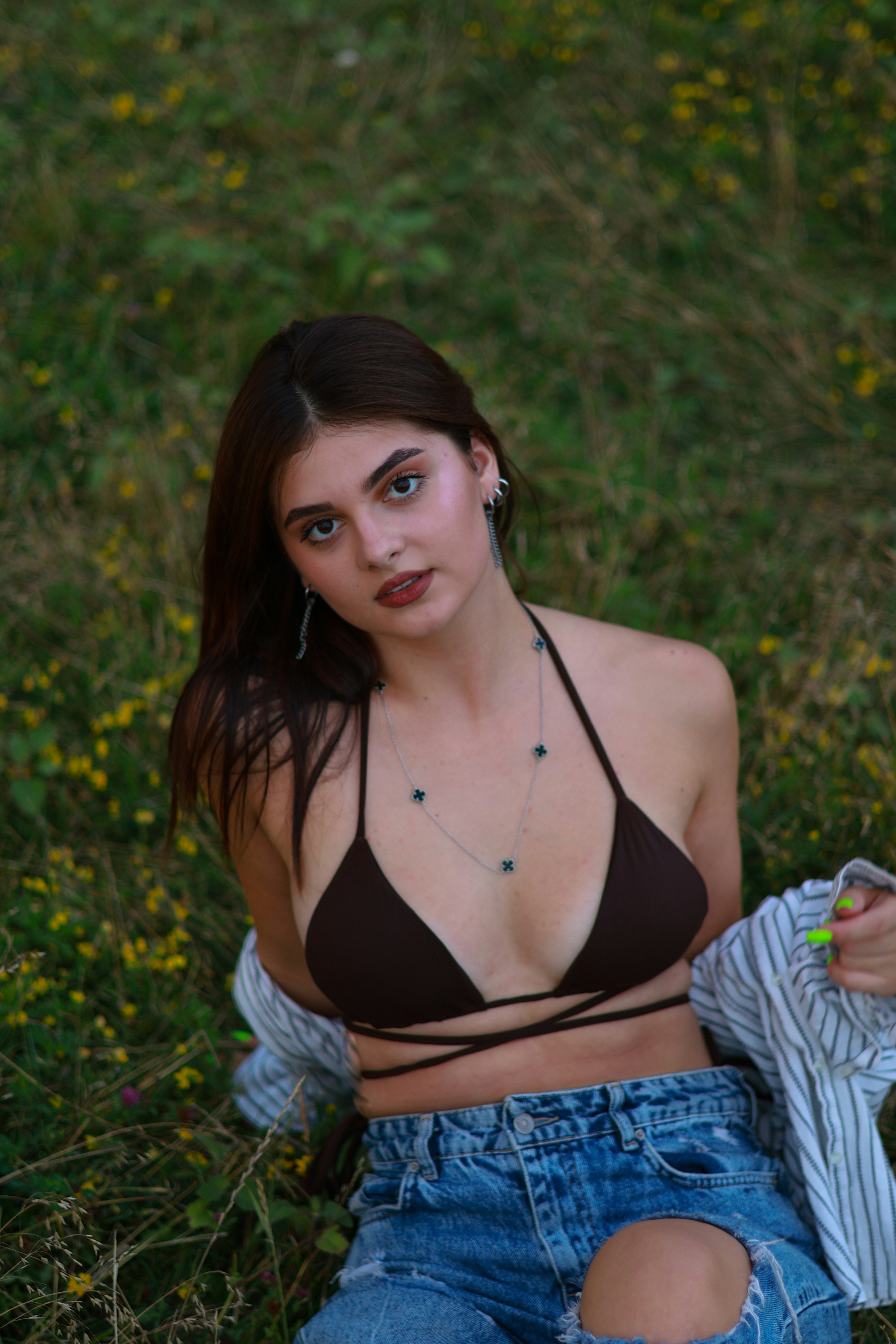 Portrait photograph of a woman in a brown bikini top and denim jeans seated amid a meadow of wildflowers with a soft green background.